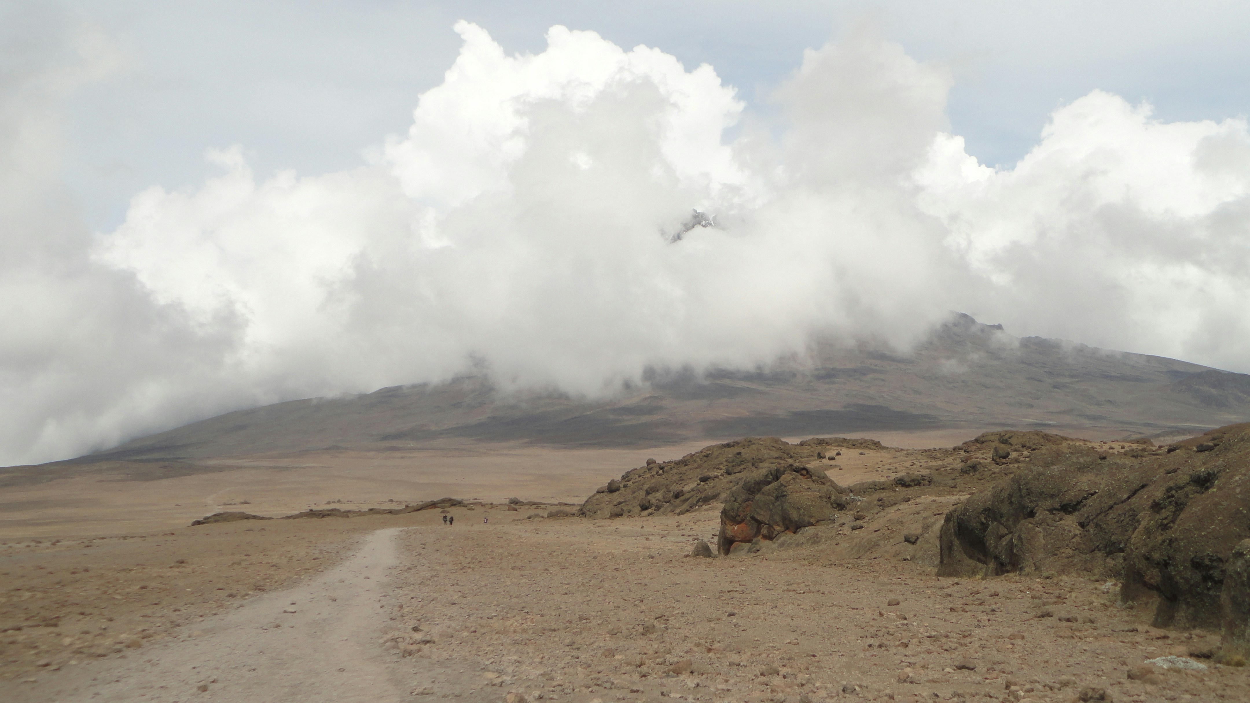 Desert trail stretches toward cloud-covered mountains, flanked by rugged rocks along a sunlit path. Landscape photograph.