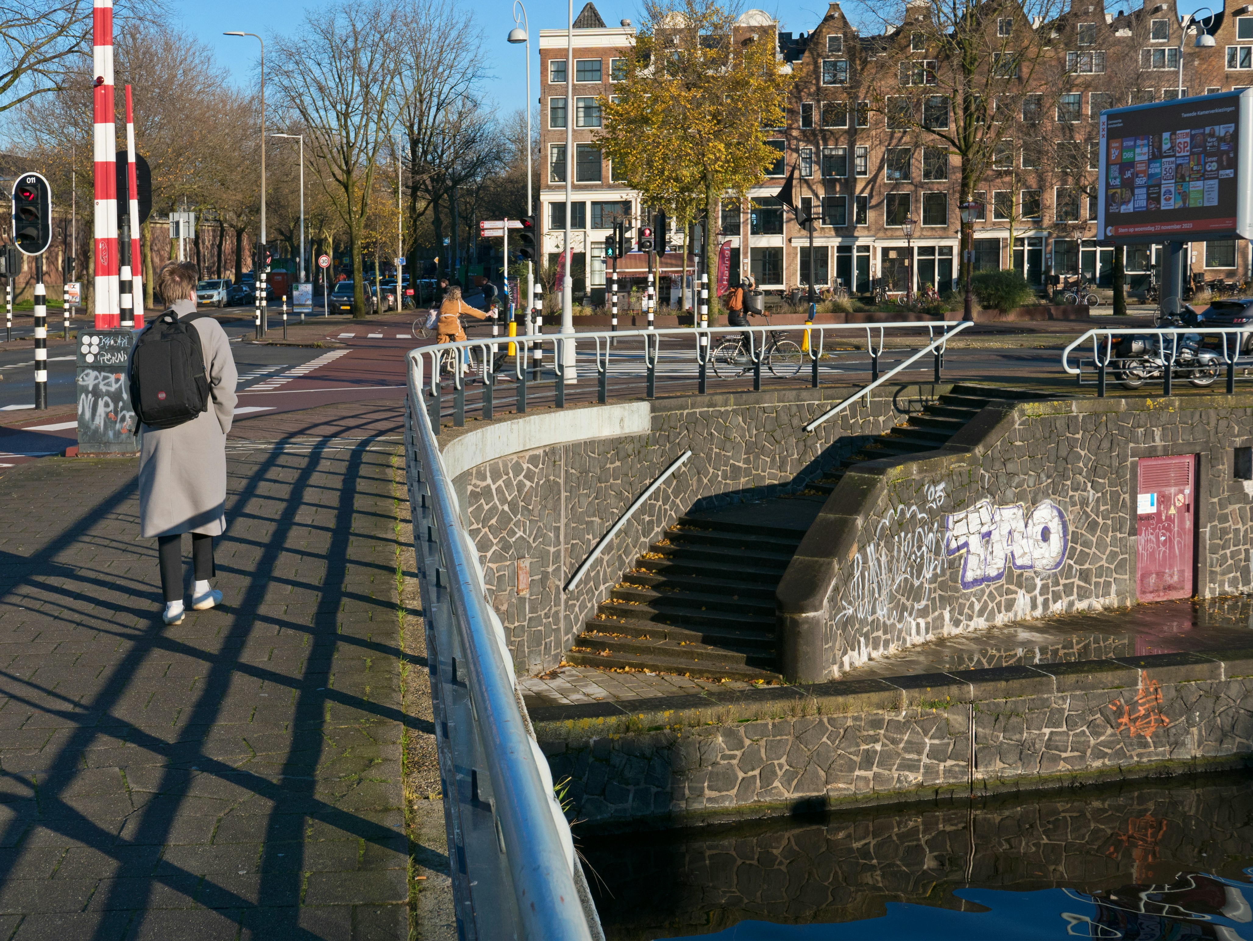 A pedestrian walks along a canal path, framed by urban architecture and autumn foliage. The scene captures the essence of city life and nature's transition.