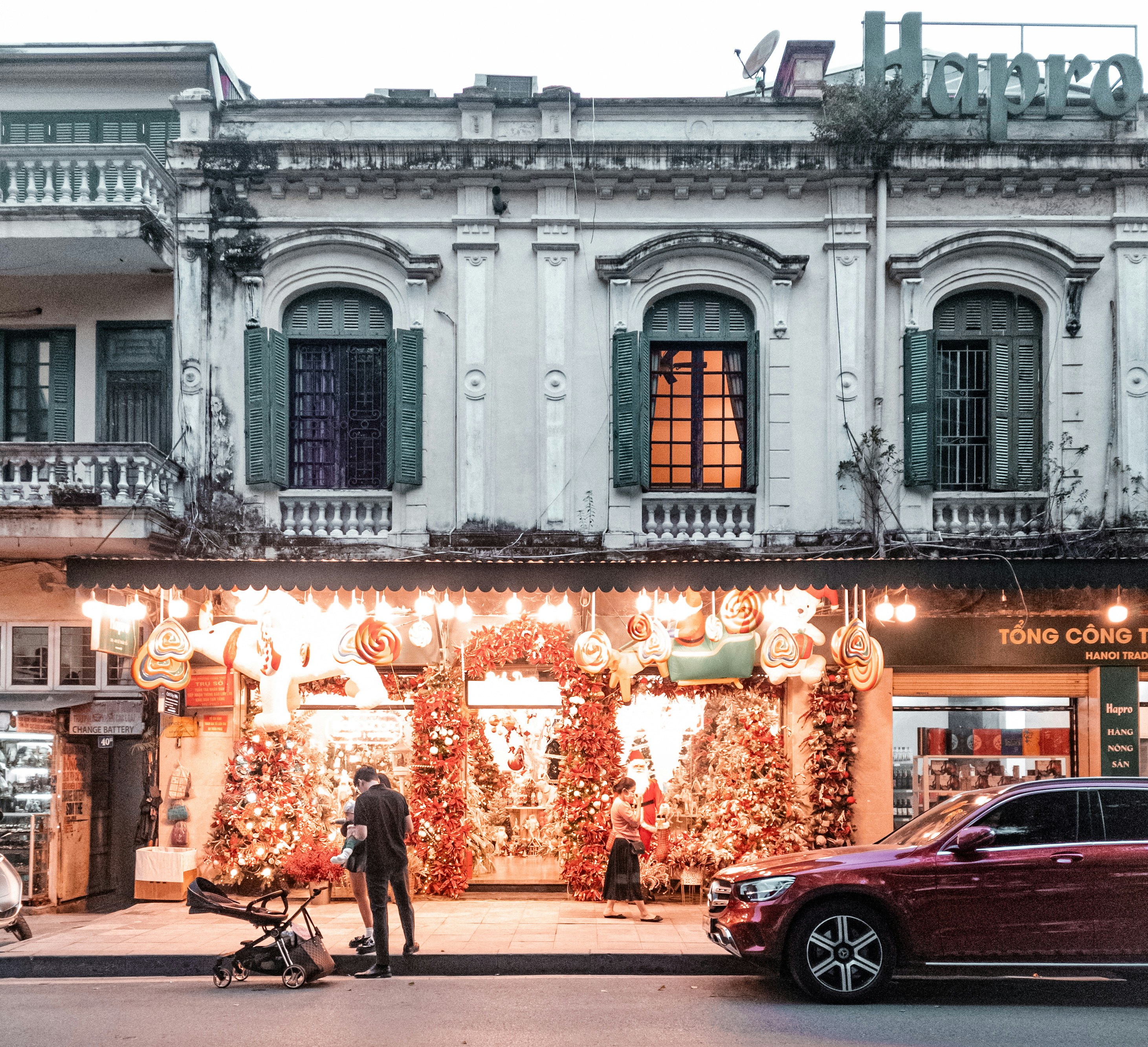 City street at dusk with a storefront festooned in Christmas lights and trees; a couple with a stroller stands before the shop while a red car sits to the right.