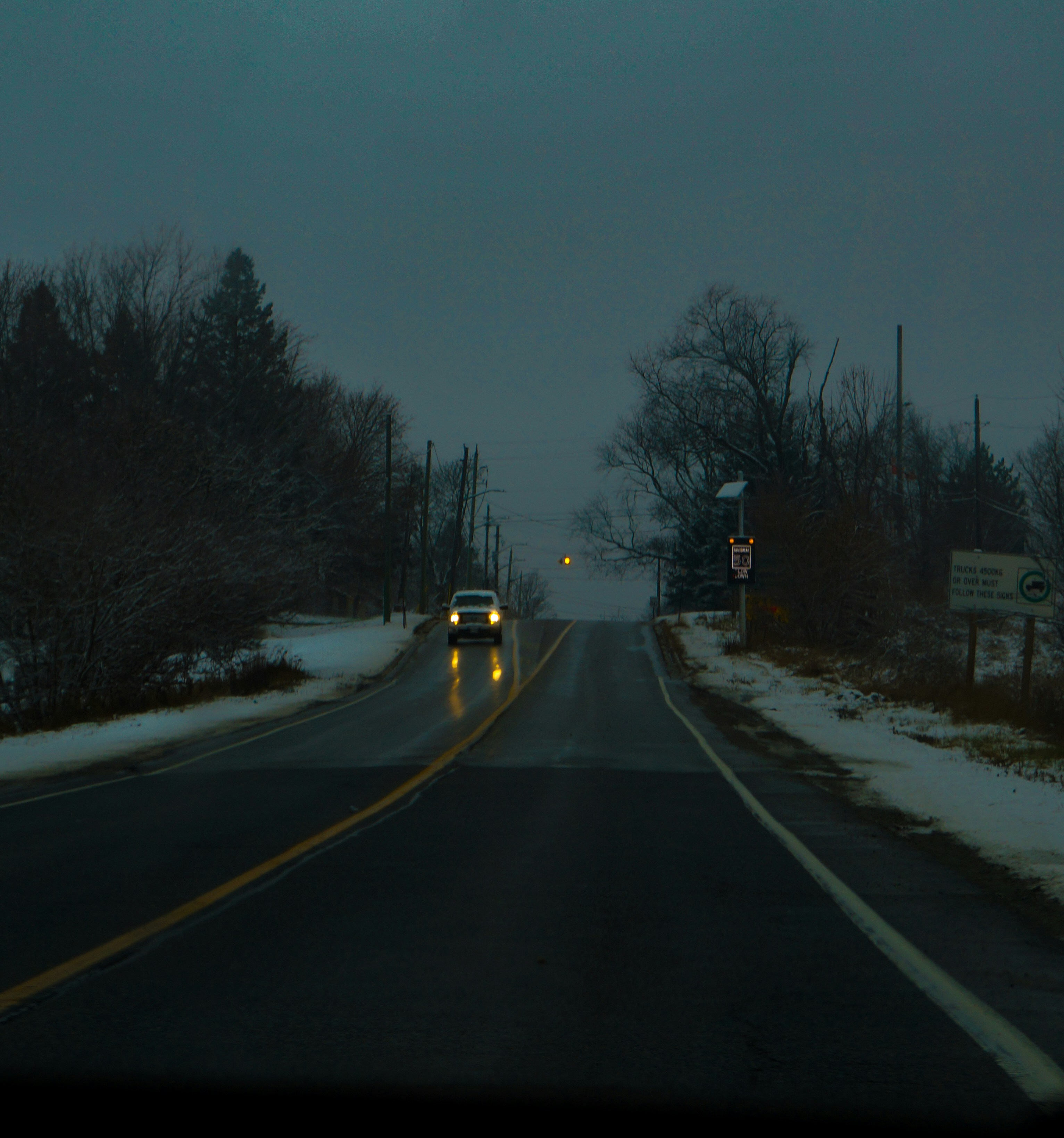 Nighttime photograph of a snow-lined rural road with a lone car approaching, its headlights reflecting on the wet asphalt.