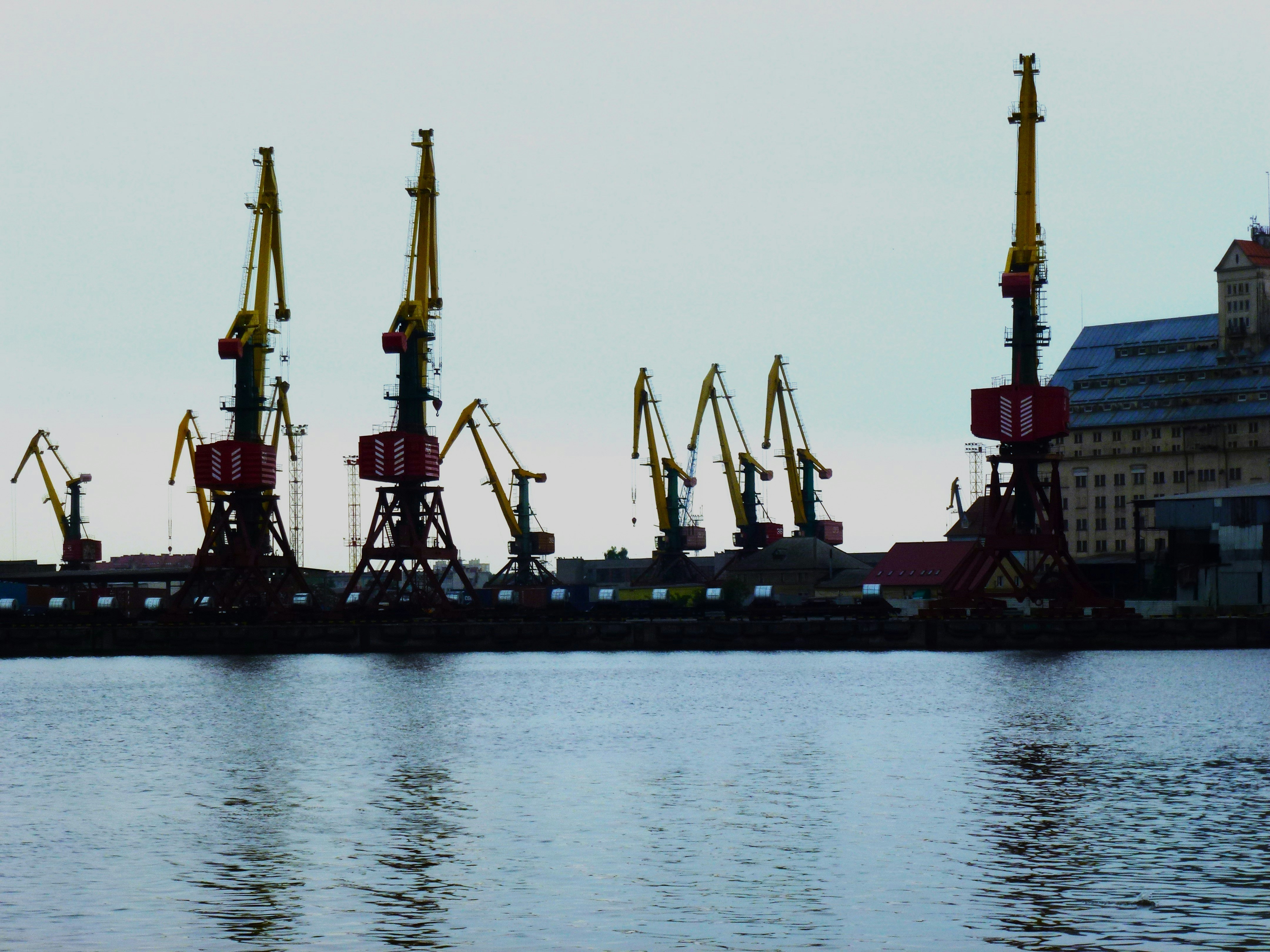 Row of harbor cranes silhouetted along the water with a distant dock and a building on the right.