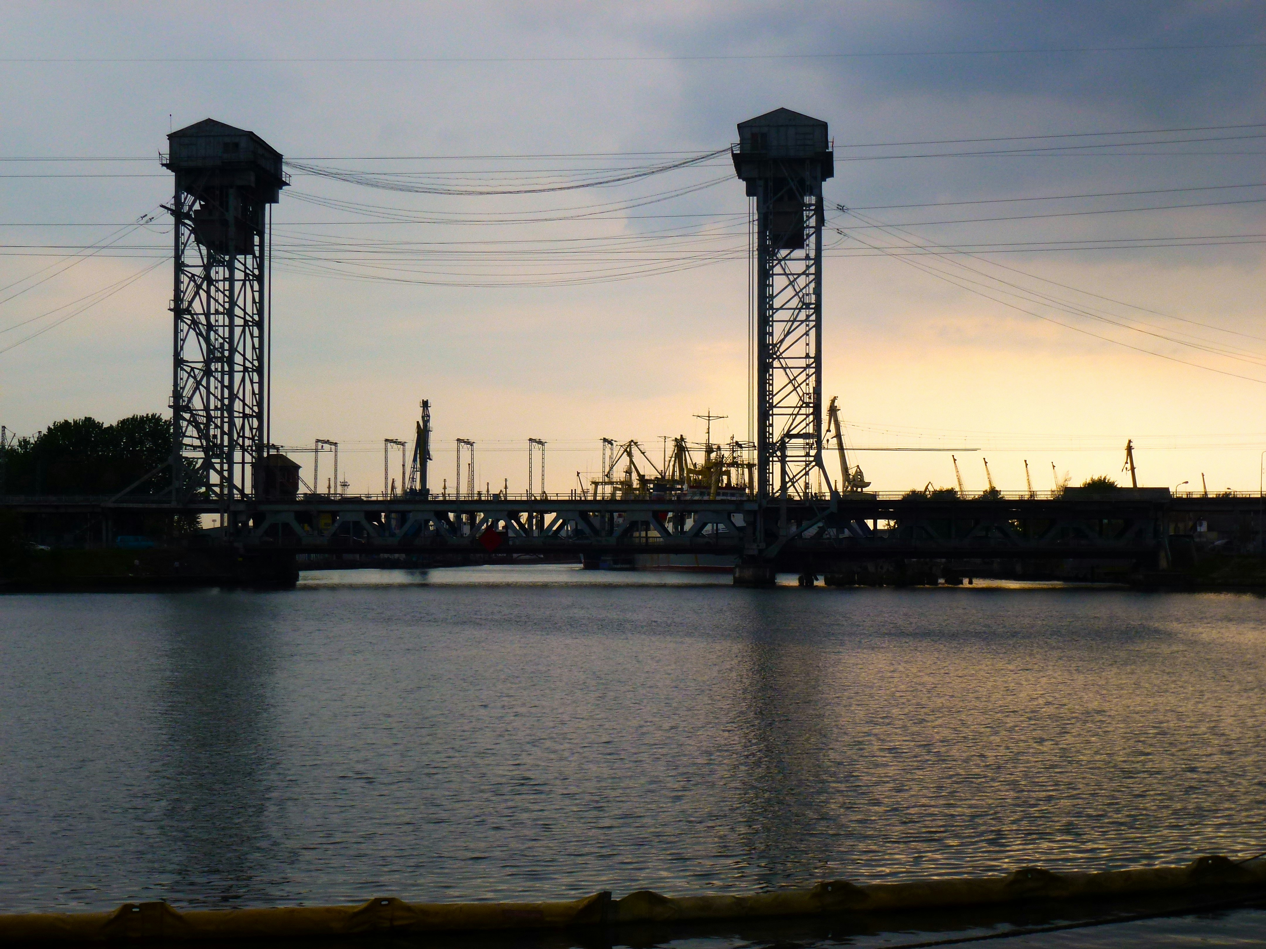 Silhouetted bridge towers rise over a calm river at sunset, with a hazy sky and distant structures lining the far bank.