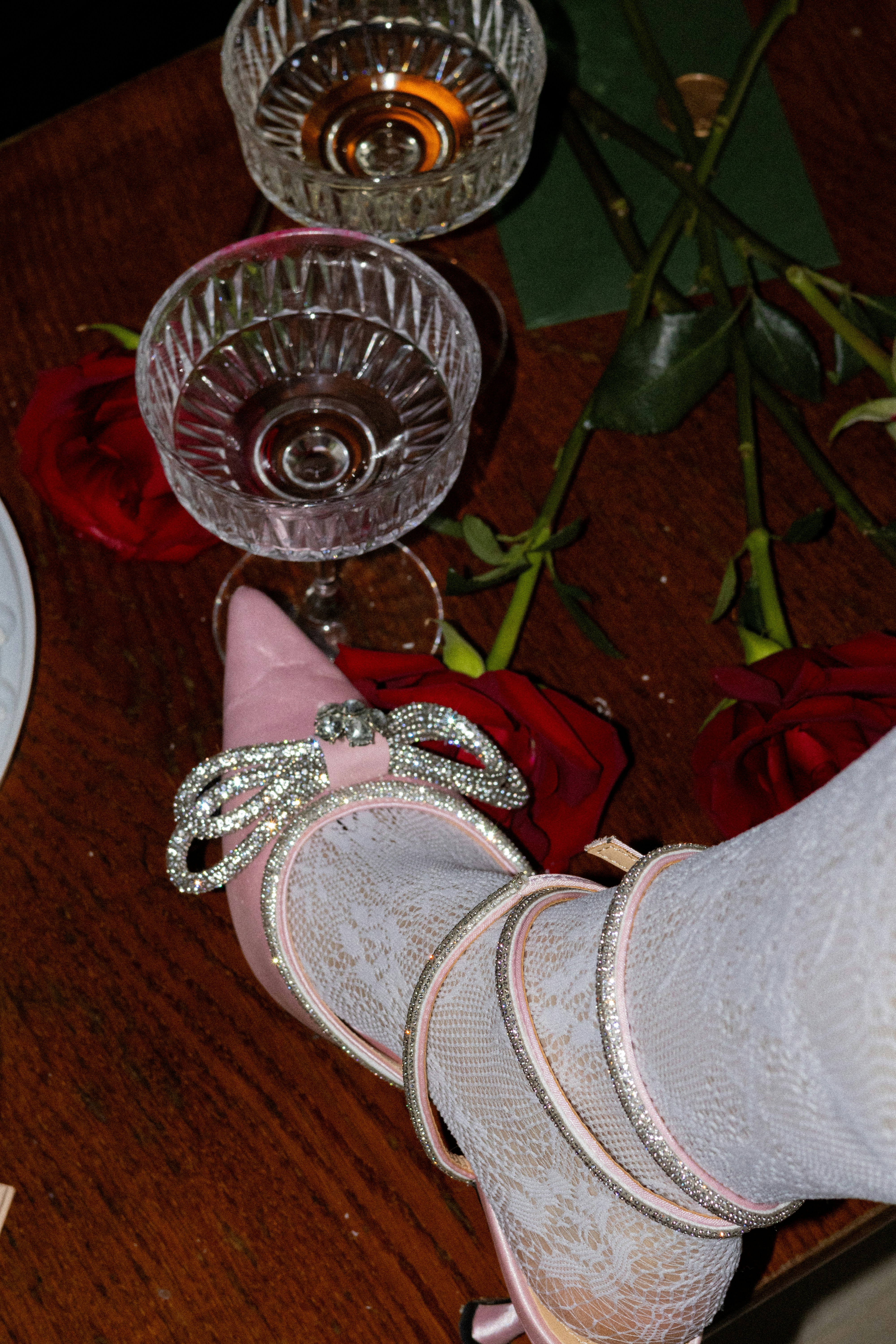 A rhinestone-embellished pink high heel rests beside a lace-clad leg on a wooden table, surrounded by red roses and carved-glass wine goblets.