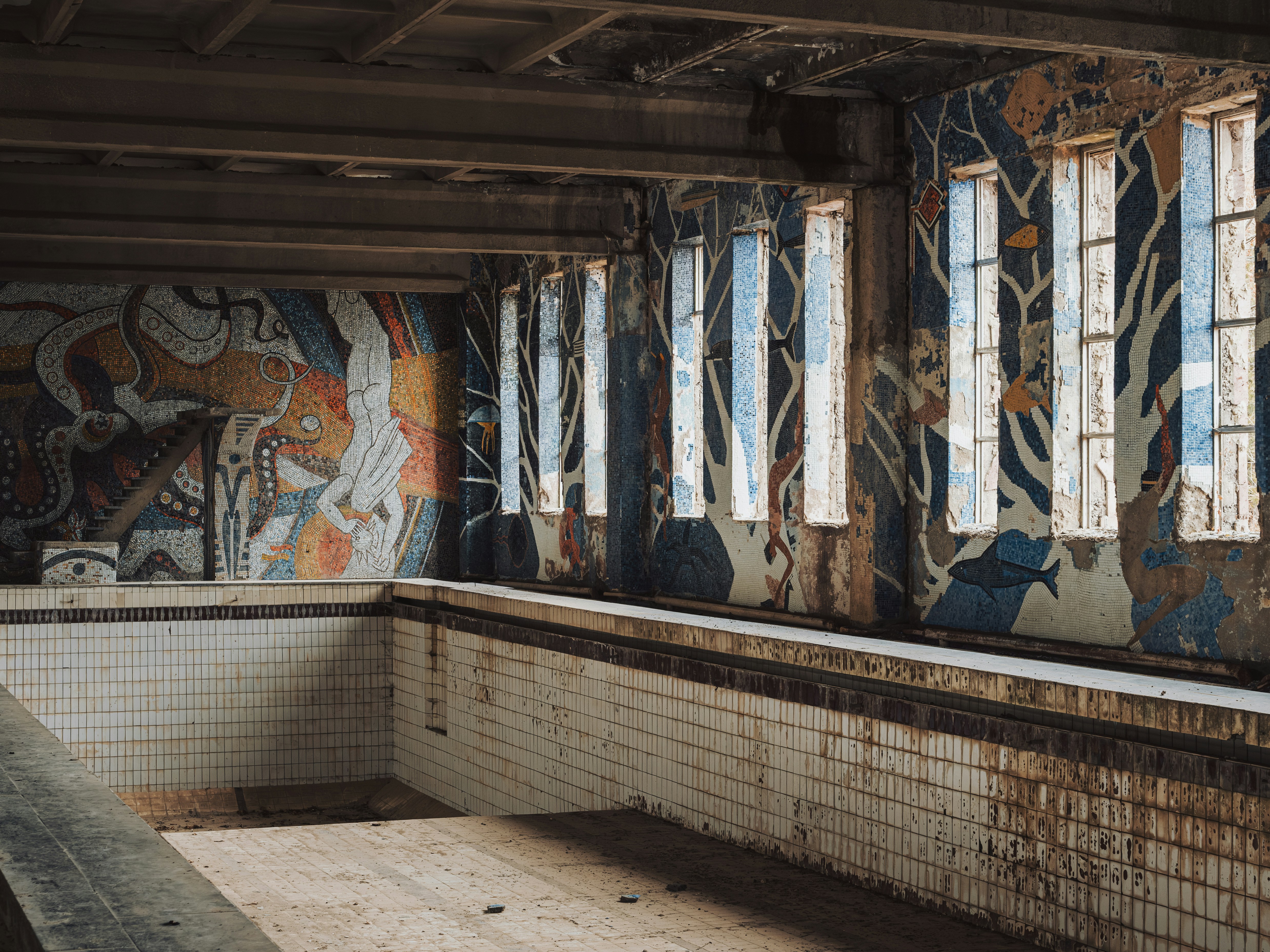 Deserted indoor pool with graffiti-covered walls and soft light streaming through tall windows.