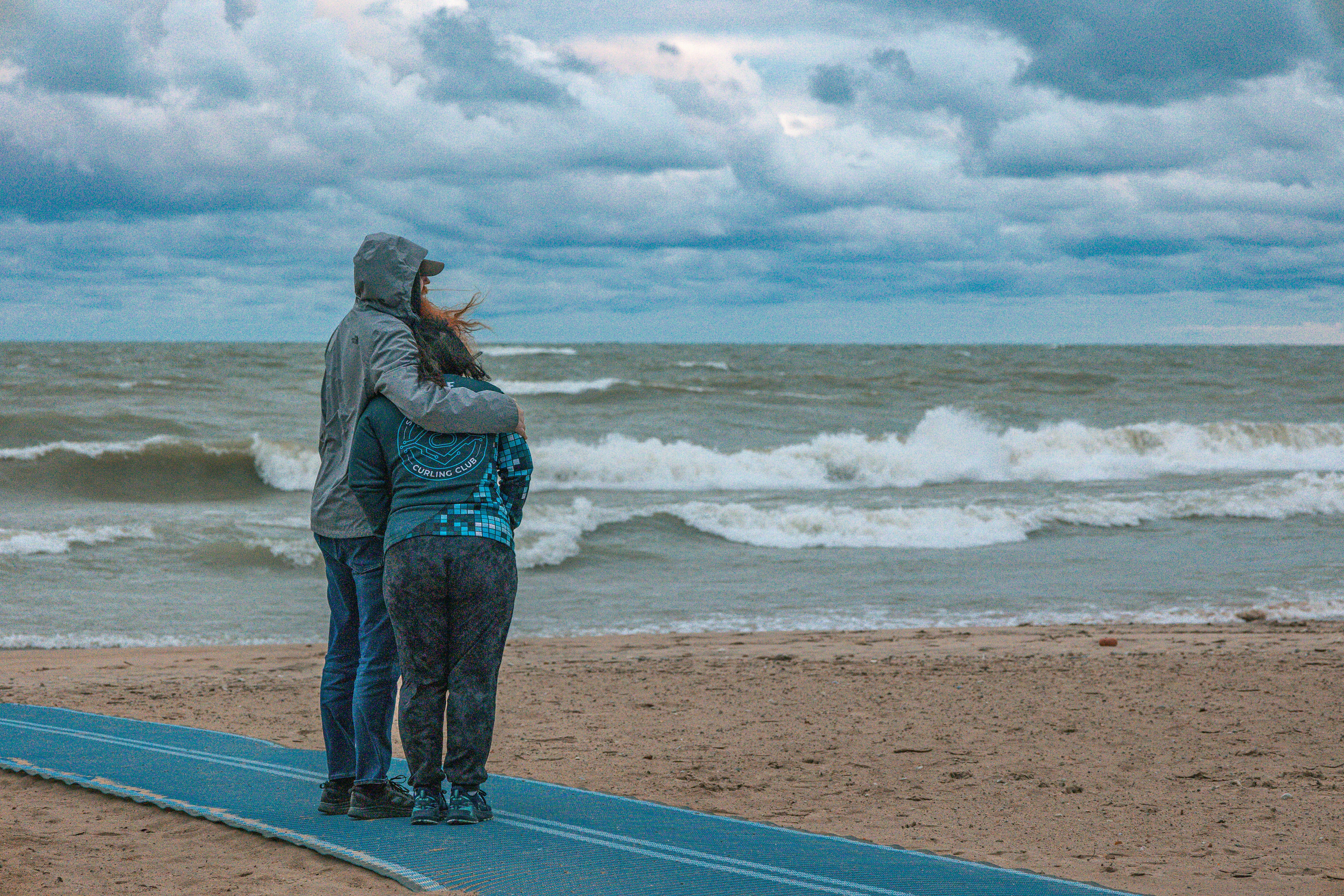 A couple stands on a sandy beach, wrapped in each other's arms, gazing at the turbulent waves under a moody, overcast sky. The image captures the contrast between the couple's serene embrace and the dynamic energy of the crashing waves, with muted blues and grays dominating the scene. The atmosphere conveys a sense of quiet intimacy against the backdrop of nature's powerful forces, making it visually striking and emotionally evocative.