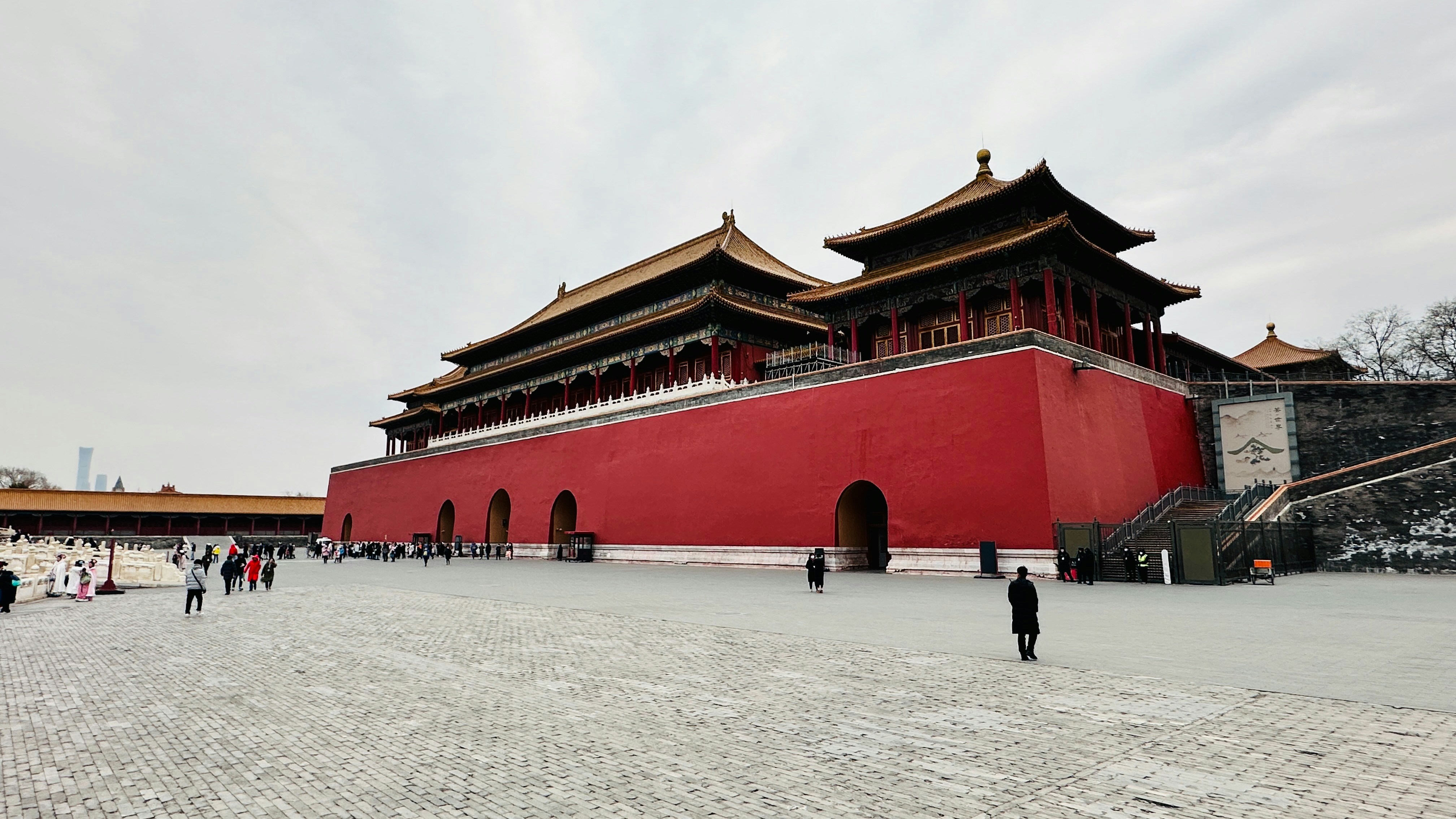 Ancient architectural marvel featuring a grand red wall and traditional rooftops, set against a cloudy sky. Visitors explore the expansive courtyard.