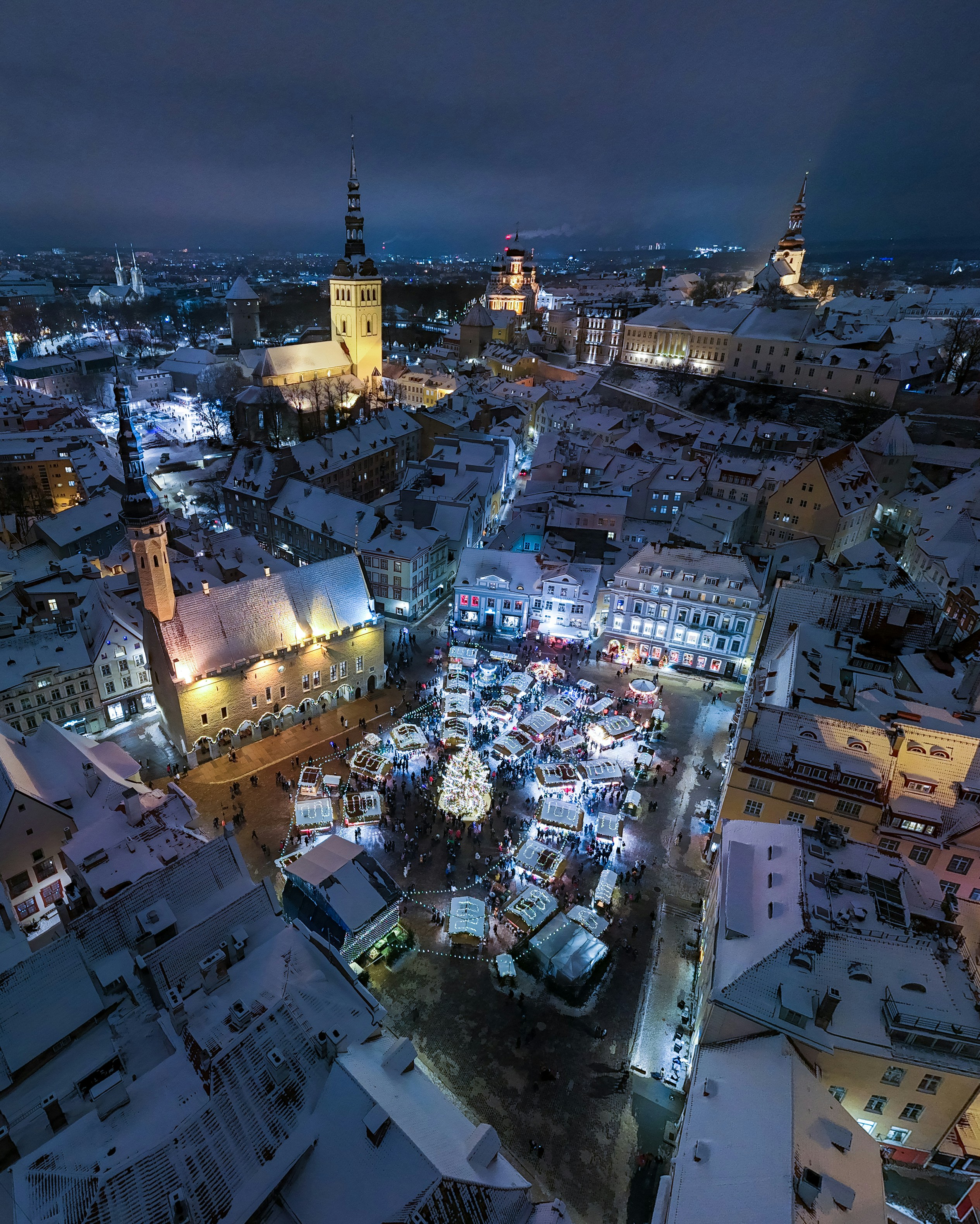 A night photograph of a snow-covered medieval town square, where a bustling Christmas market glows with string lights amid winding rooftops and historic towers.