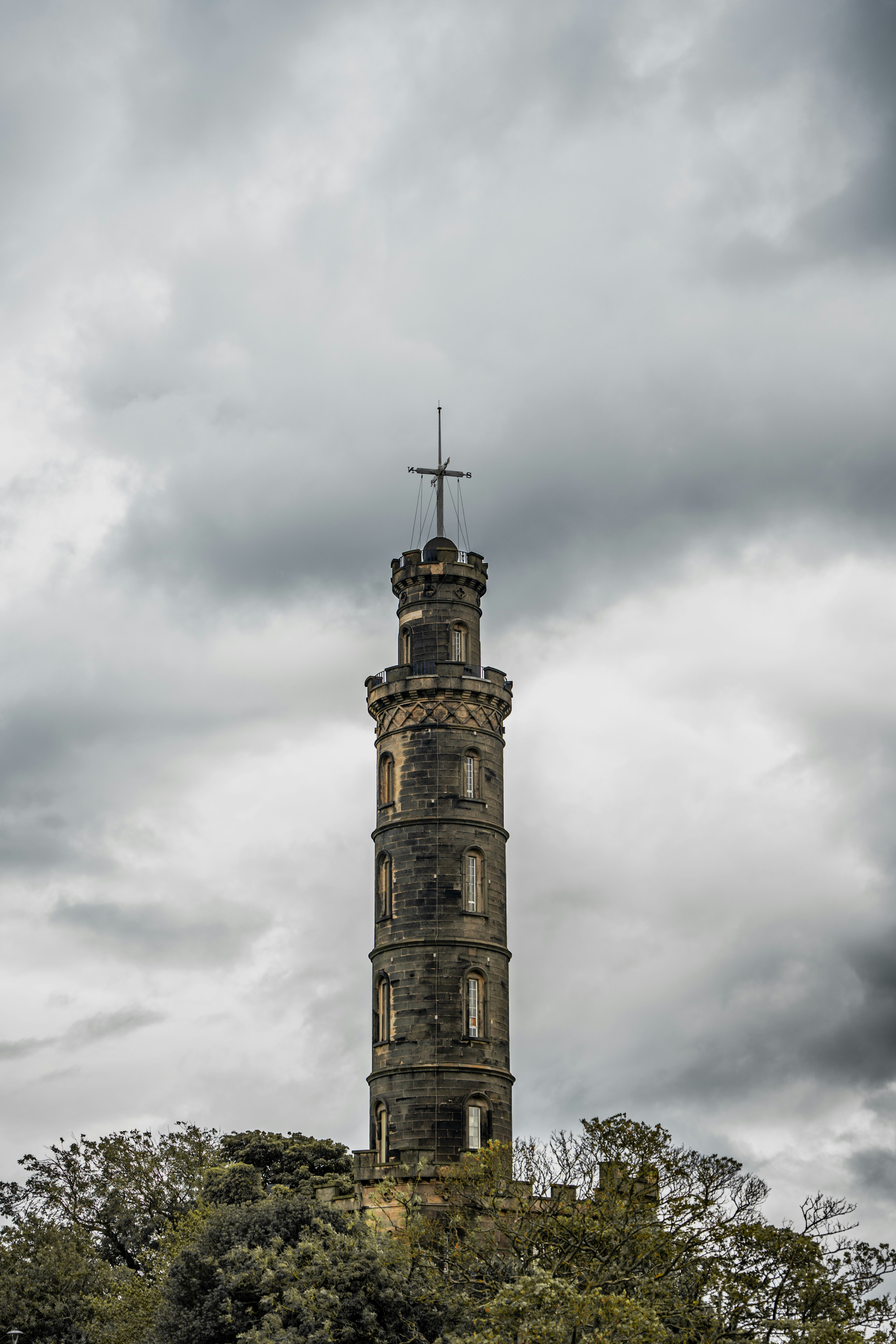 A tall, historic tower rises dramatically against a backdrop of dark, swirling clouds, surrounded by lush greenery.