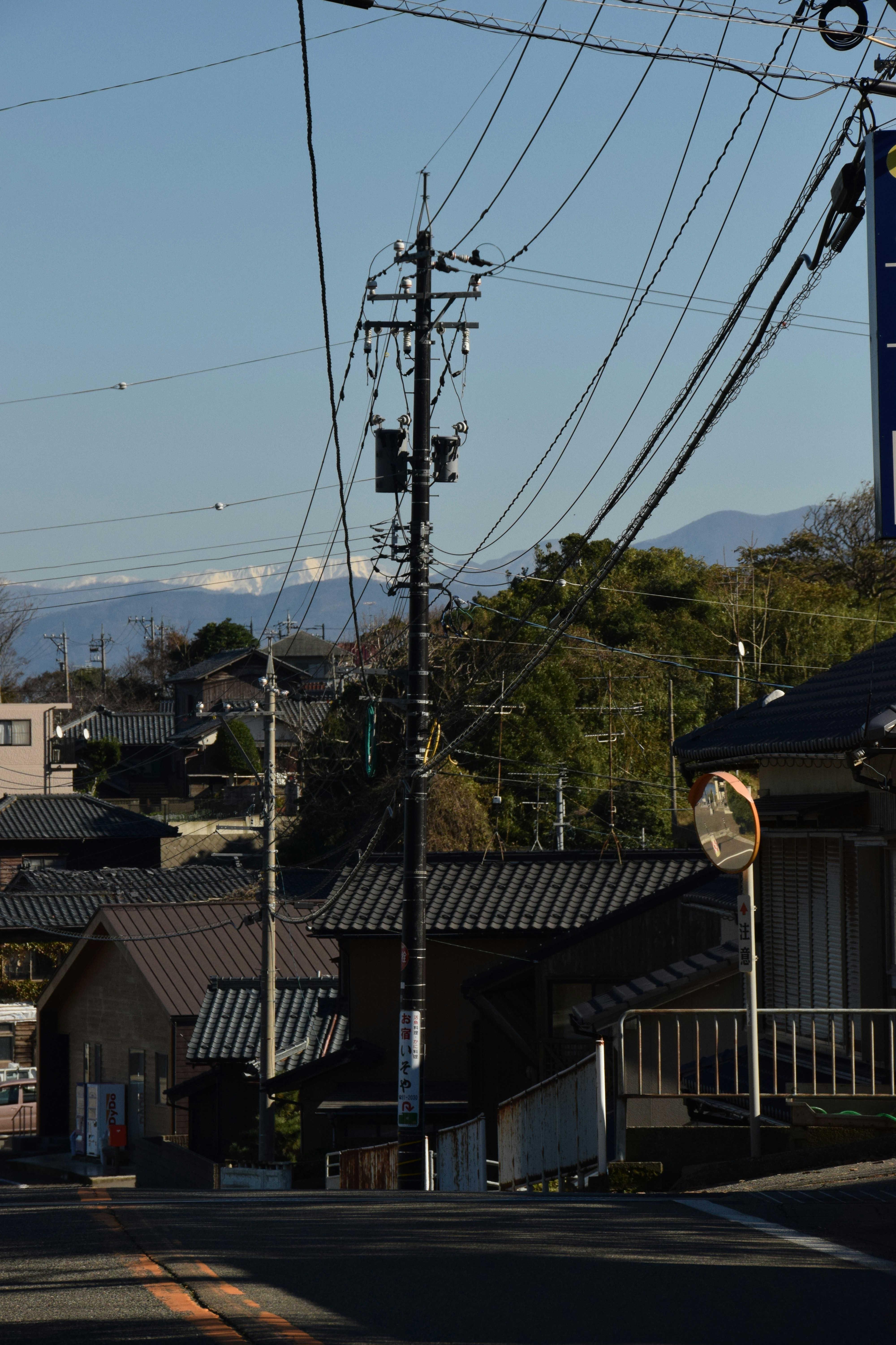 A quiet street scene showcasing traditional Japanese houses interspersed with utility poles and power lines against a mountainous backdrop. The composition reflects the blend of nature and urban life.