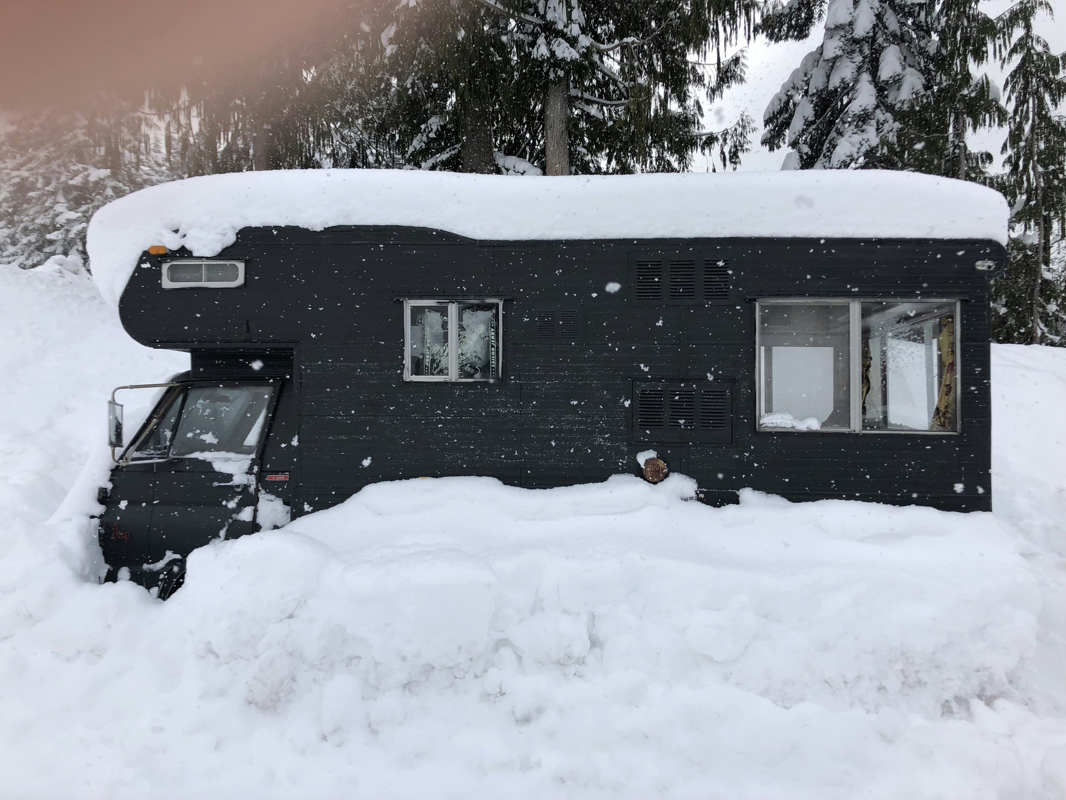 A snow-covered camper partially buried in a winter landscape, surrounded by towering evergreen trees. The scene captures the essence of solitude and resilience in harsh weather conditions.