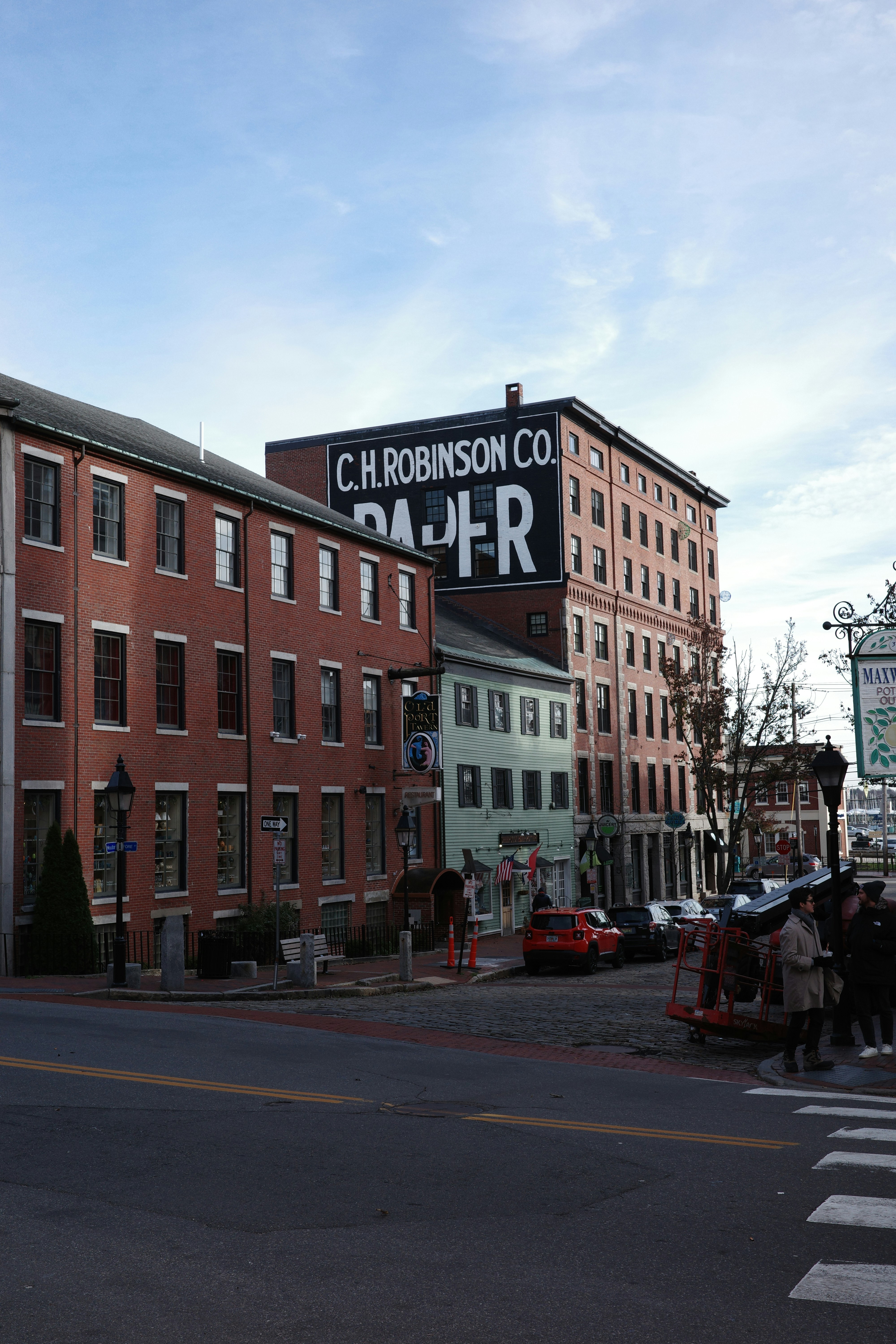 Historic brick buildings line a cobblestone street, with a large vintage advertisement for C.H. Robinson Co. prominently displayed. The scene captures the charm of a bygone era in a bustling urban setting.