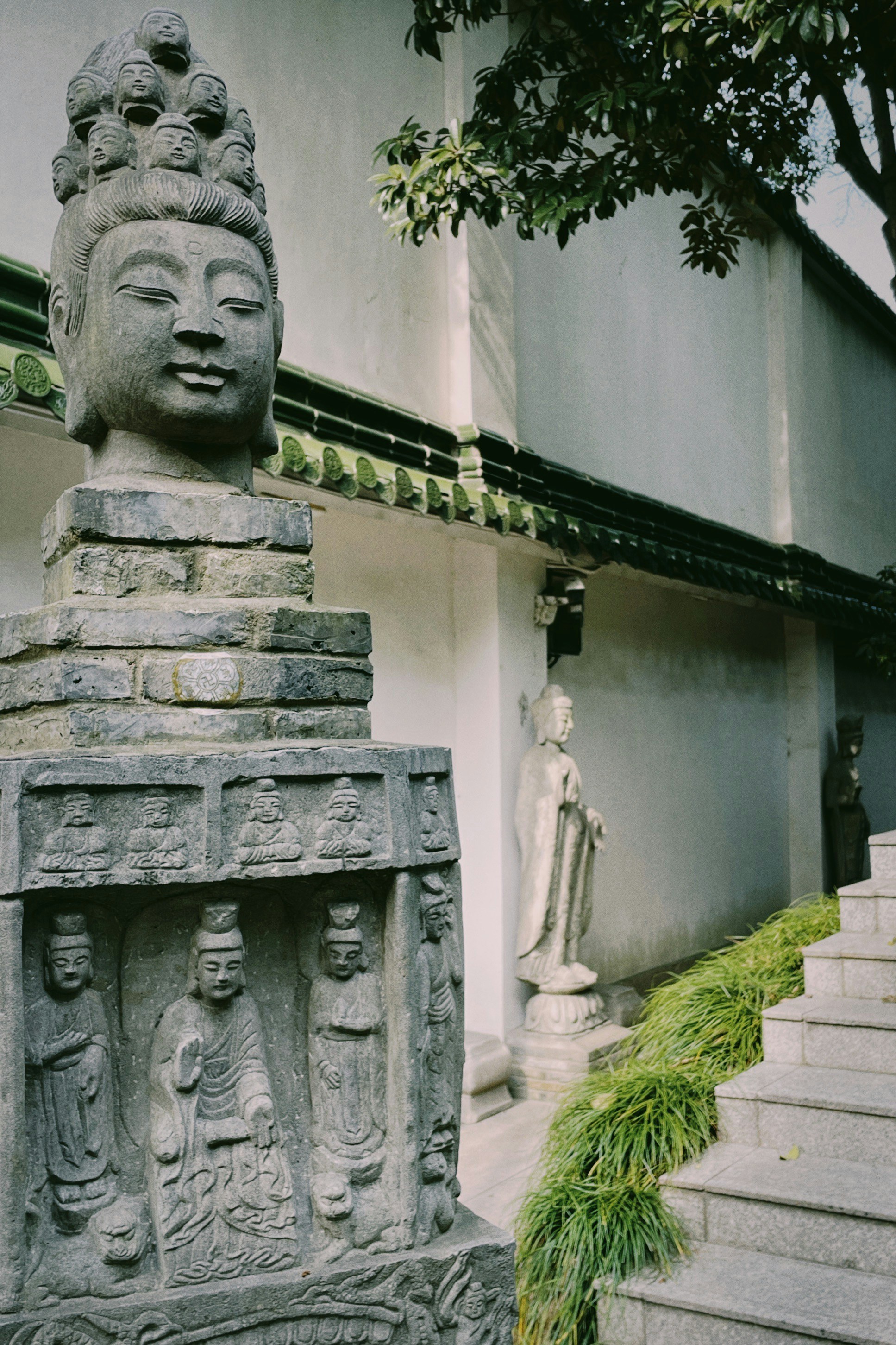 A large carved stone statue anchors a quiet courtyard, flanked by relief panels and smaller figures along a stepped pedestal. The scene conveys a tranquil, ancient atmosphere beside a white wall and greenery.