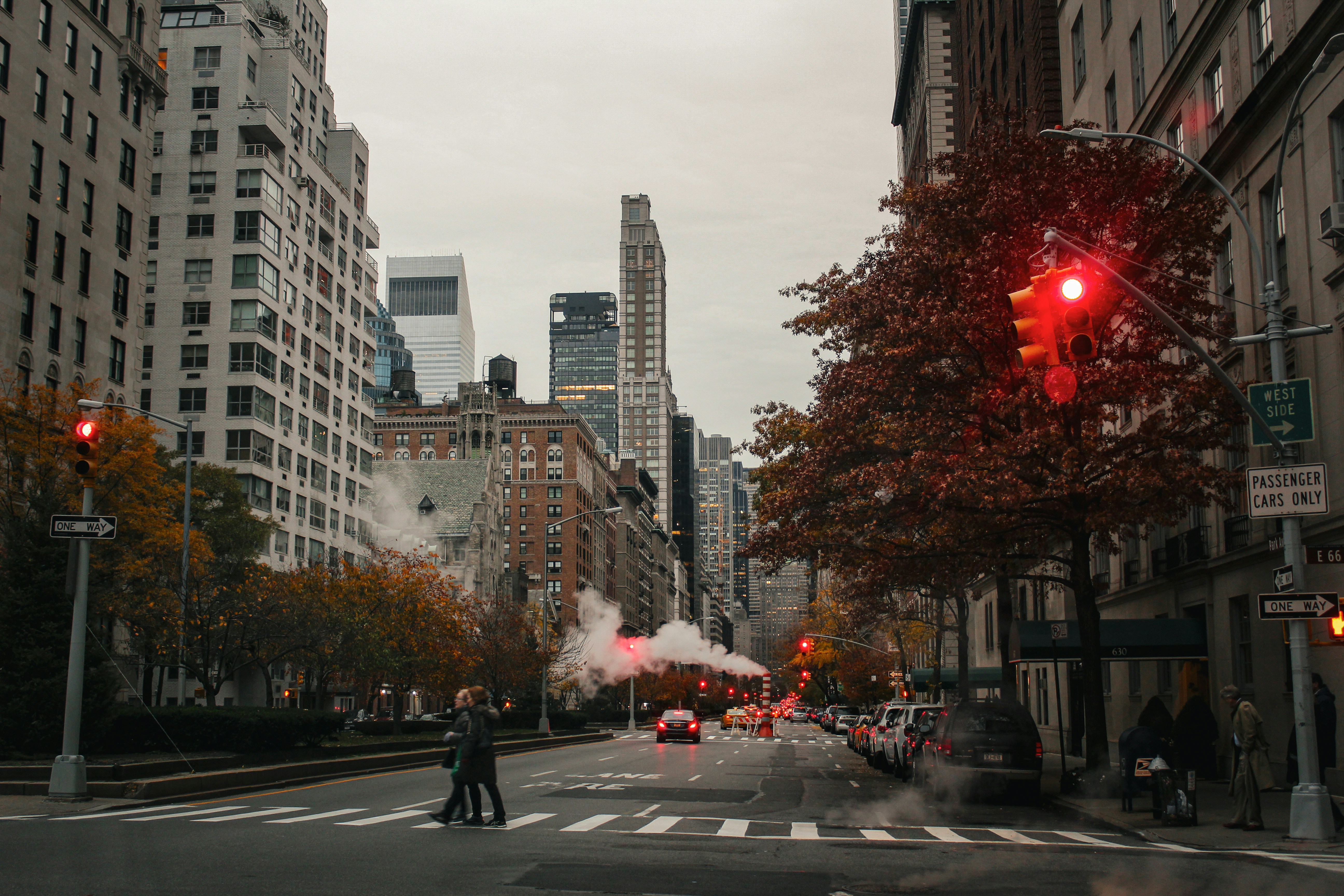 Busy city street with pedestrians crossing, surrounded by towering buildings and steam rising from manholes. Red traffic light adds to the urban atmosphere.