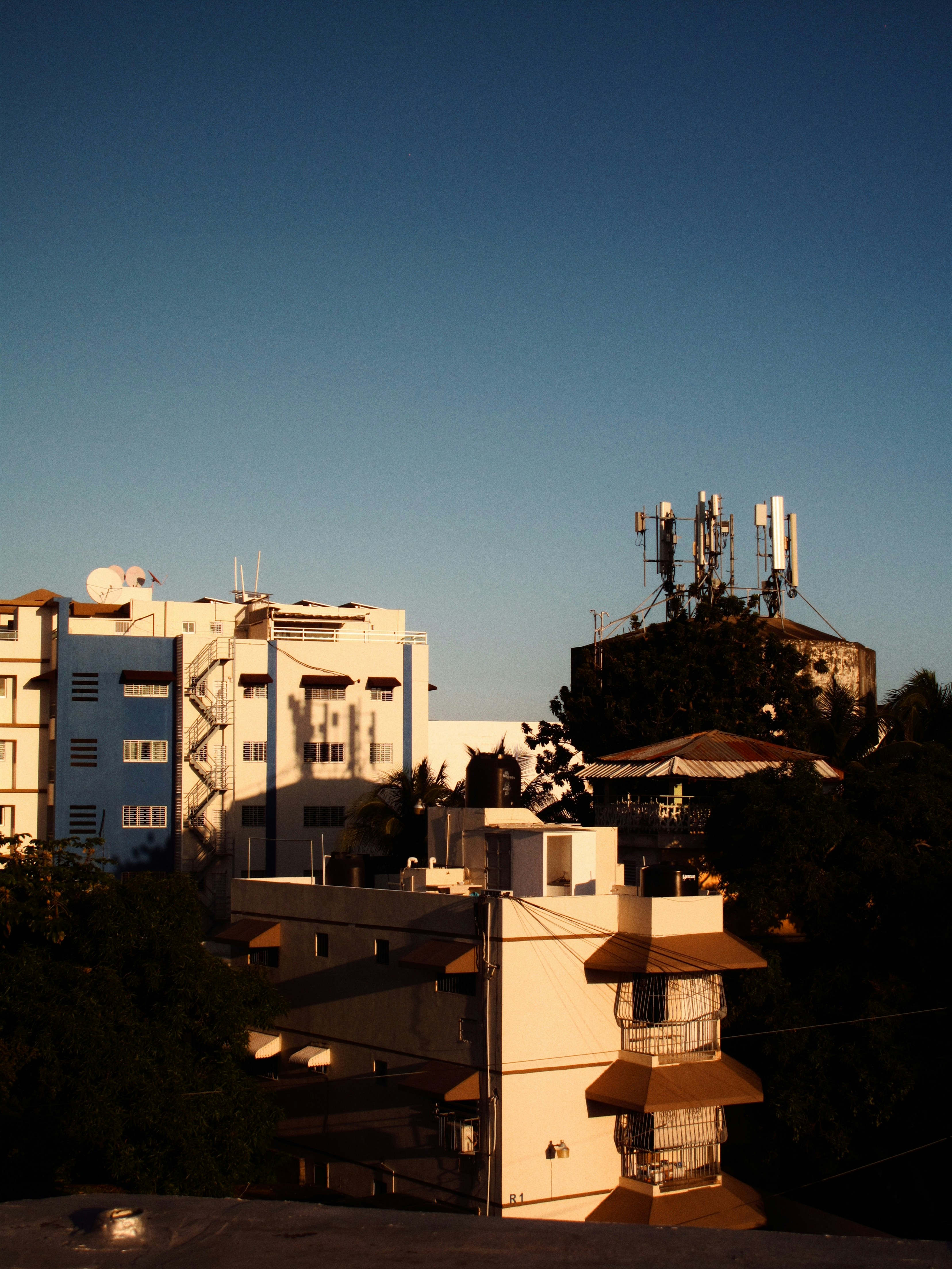 Cityscape at golden hour, foreground featuring a blue apartment block and a rooftop communications tower against a smooth gradient sky.