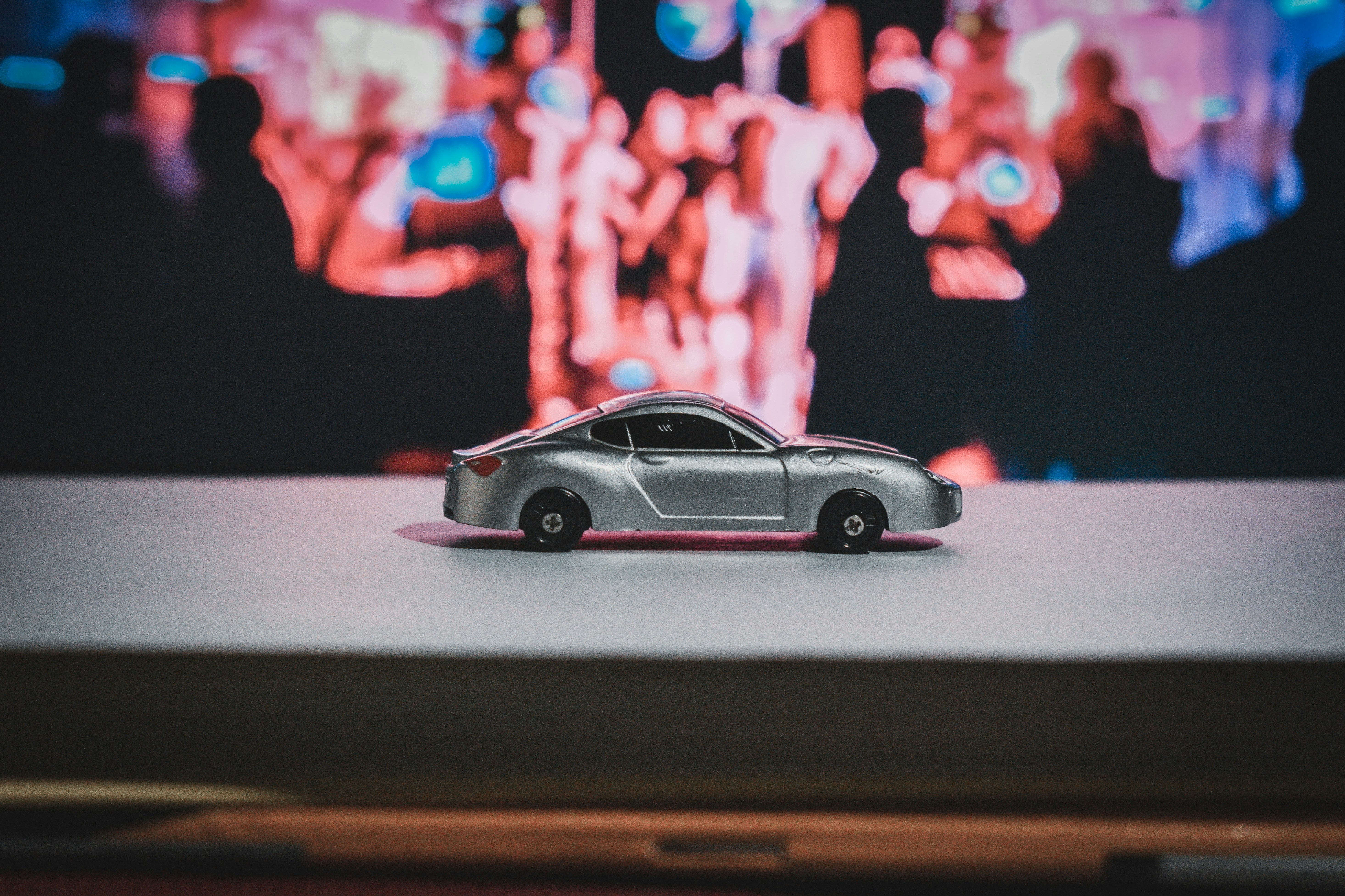 A sleek silver toy car positioned on a flat surface, with a vibrant, blurred background of colorful lights and figures.