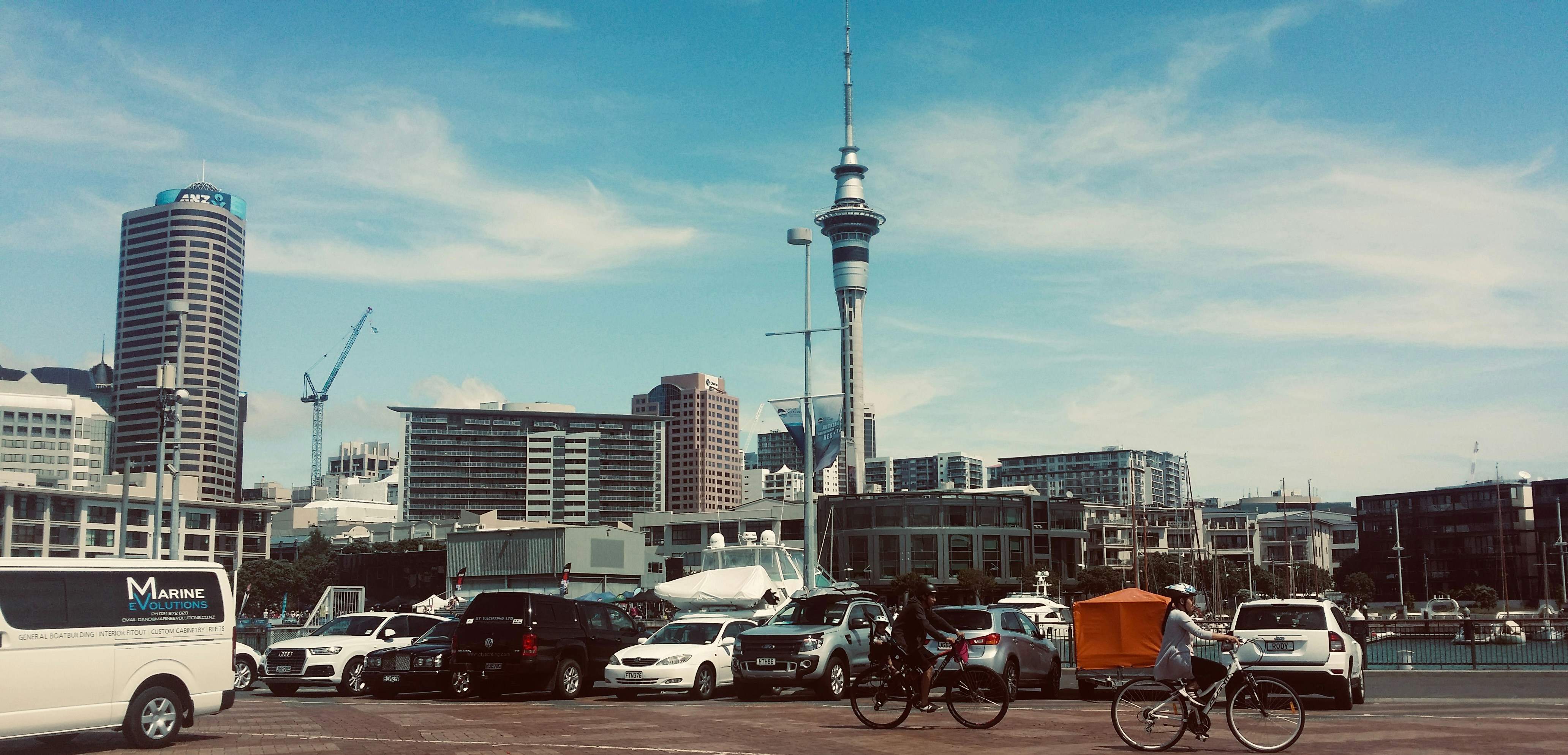 Sky Tower dominates the skyline against a clear blue sky. A row of parked cars and cyclists fills the foreground of the urban scene.
