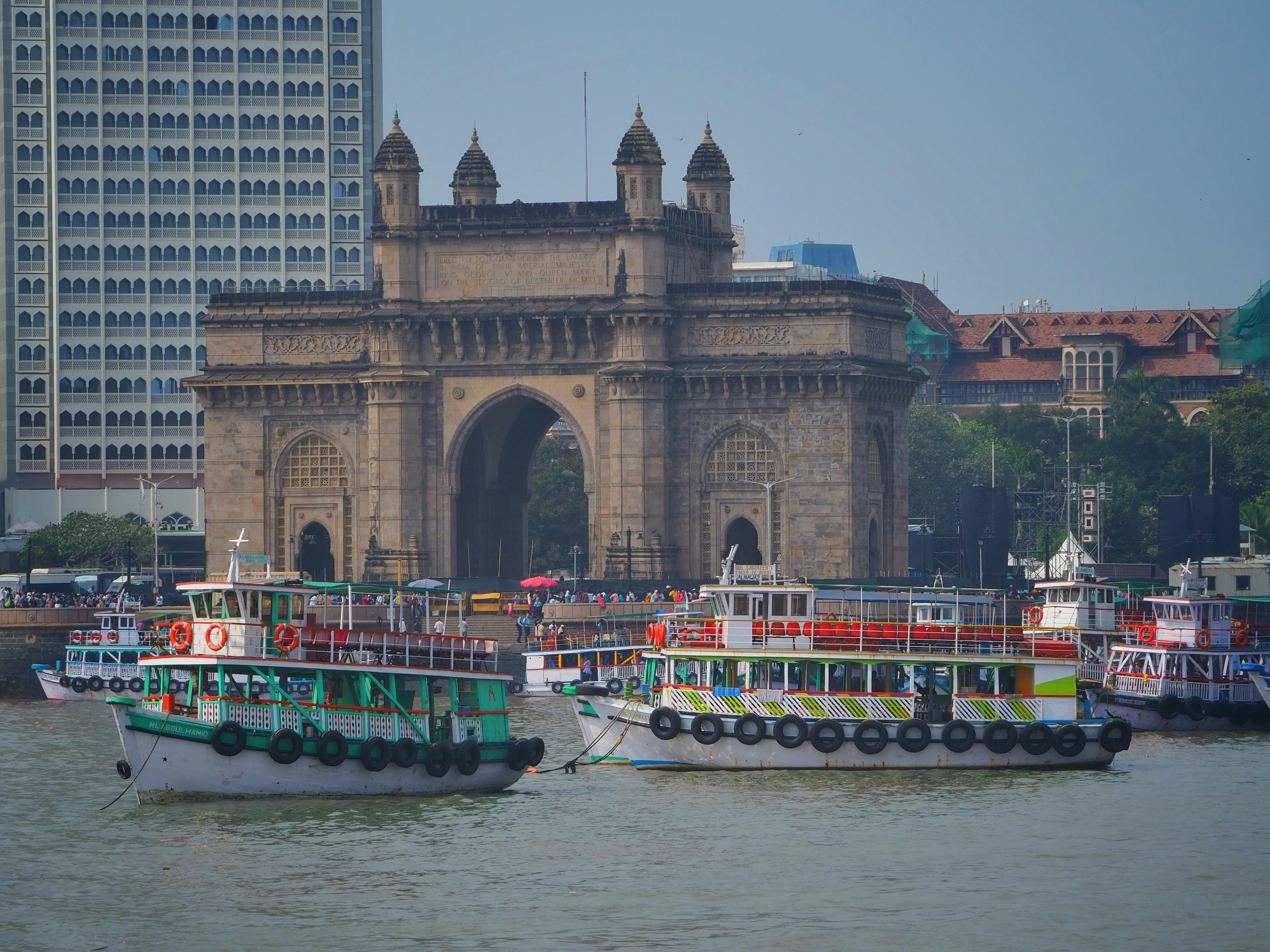 Colorful boats navigating the waters in front of the Gateway of India, a historic archway in Mumbai, with modern buildings in the background.