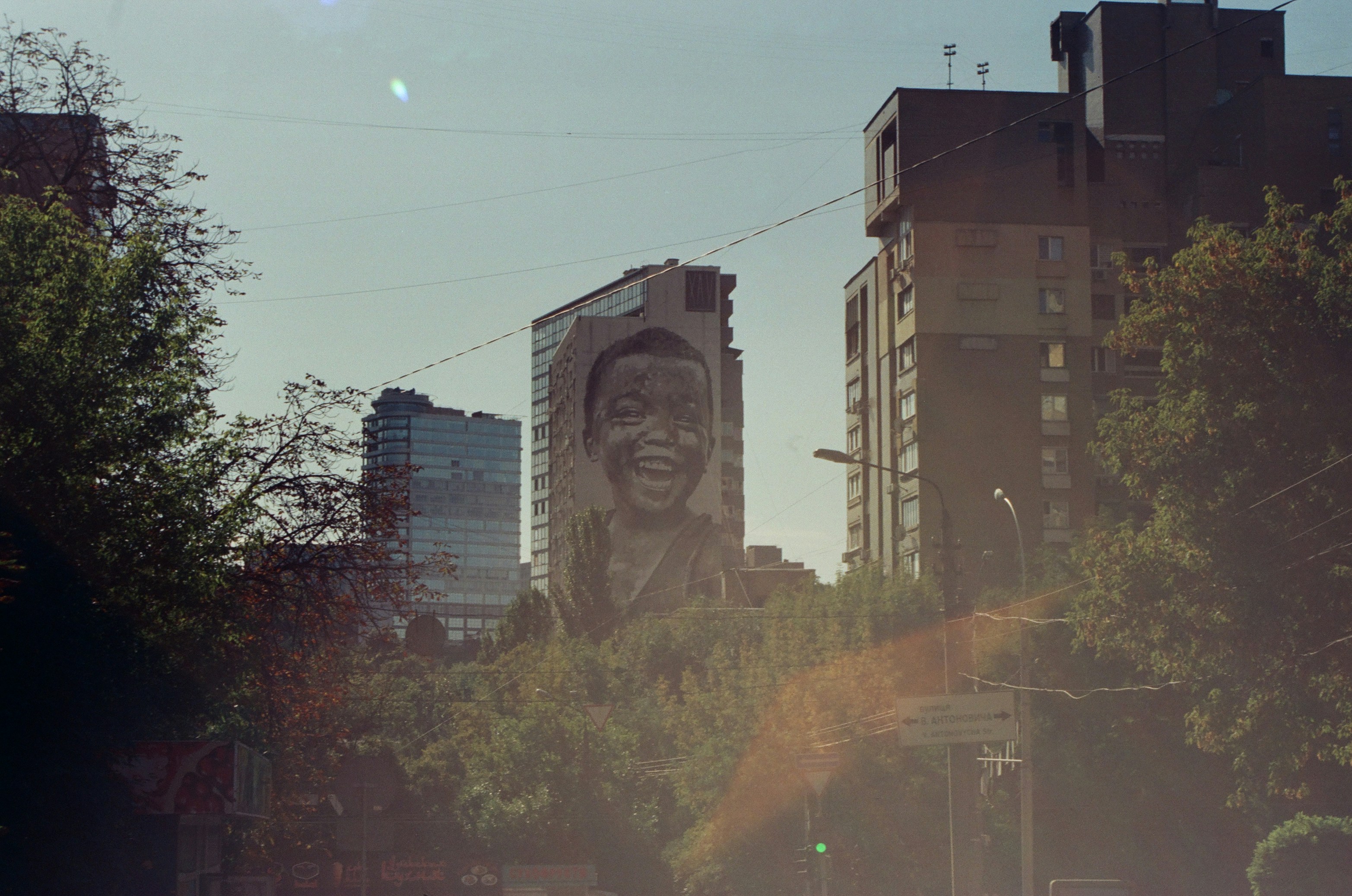 City street with a towering mural painting of a smiling face on a building, framed by trees and urban high-rises.