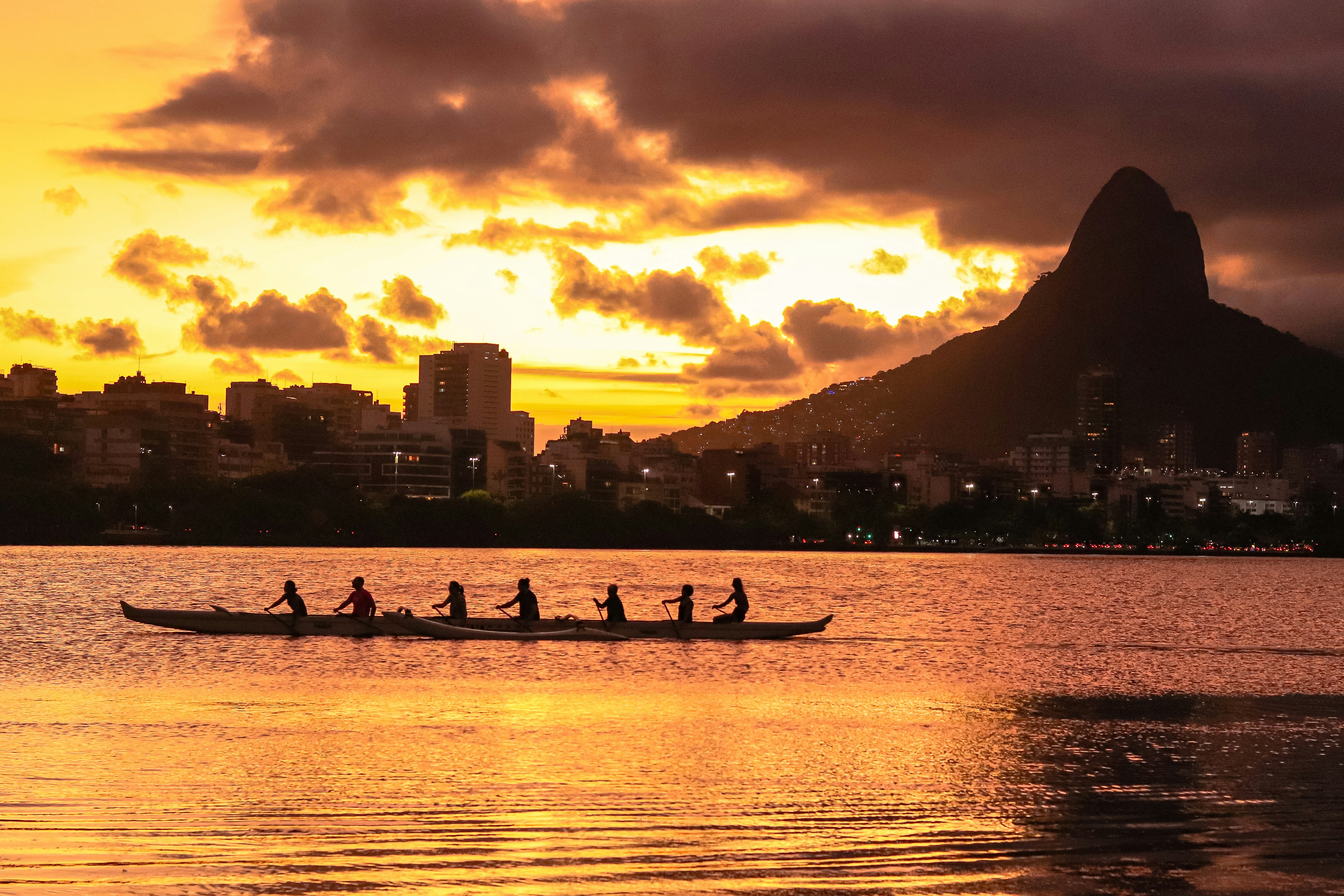 Rowers in a long boat glide across a tranquil body of water at sunset with a mountain silhouette in the background.