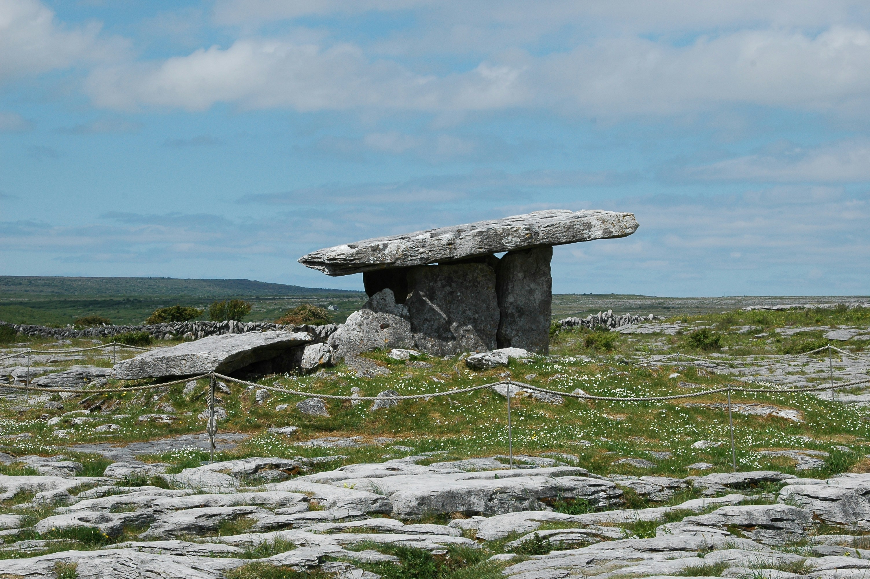 Neolithic dolmen standing resiliently on a rocky landscape under a blue sky, surrounded by lush greenery.