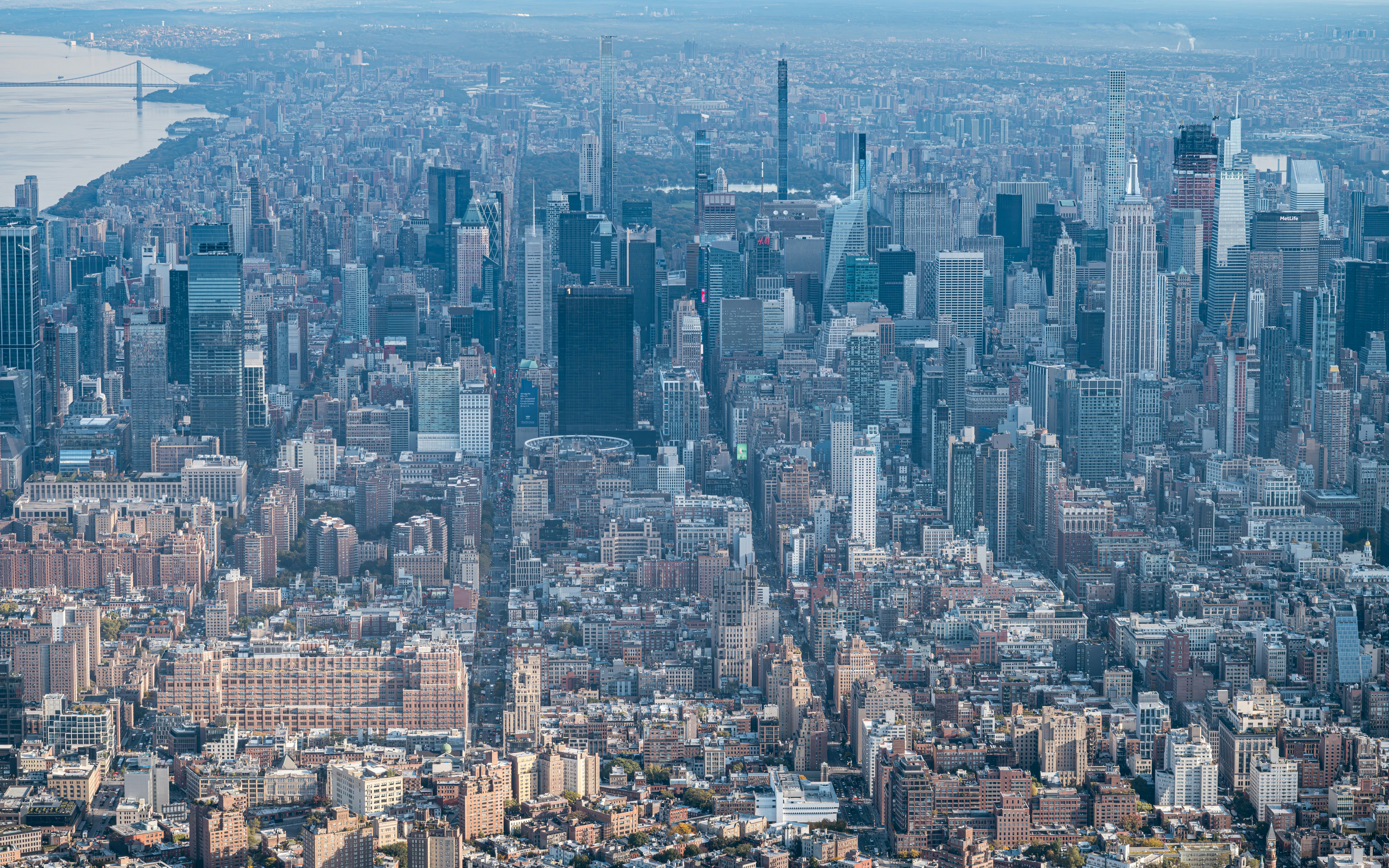 Aerial cityscape of Manhattan's dense skyline viewed from above, with the river on the left and a sea of glass-clad high-rises filling the frame. Emphasizes geometric patterns and depth across the urban expanse.