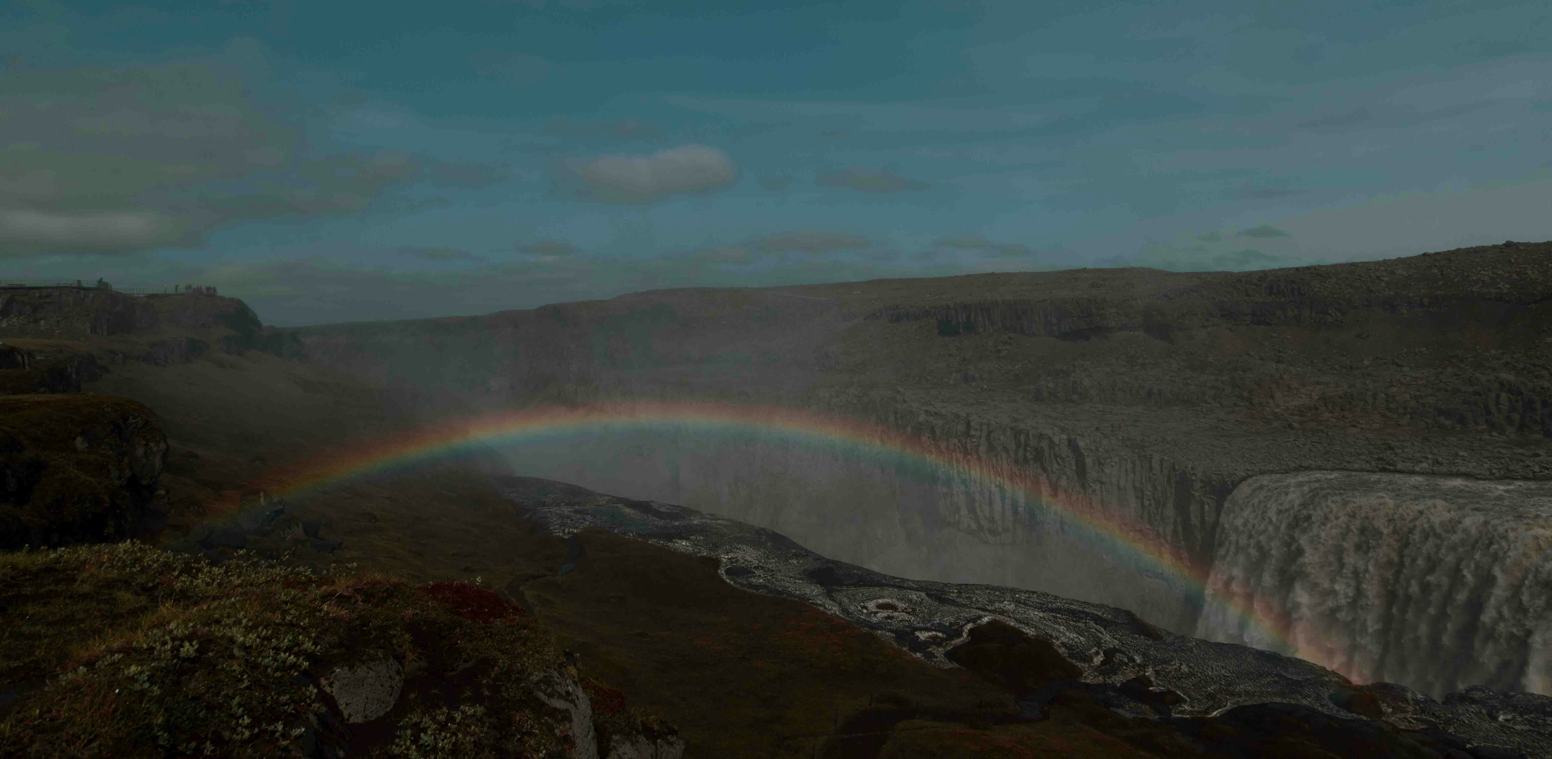 This image captures the ethereal beauty of a rainbow arcing over the mist of Dettifoss, one of Iceland's most powerful waterfalls. The lush green foreground and rugged canyon walls frame the falls' raw power.