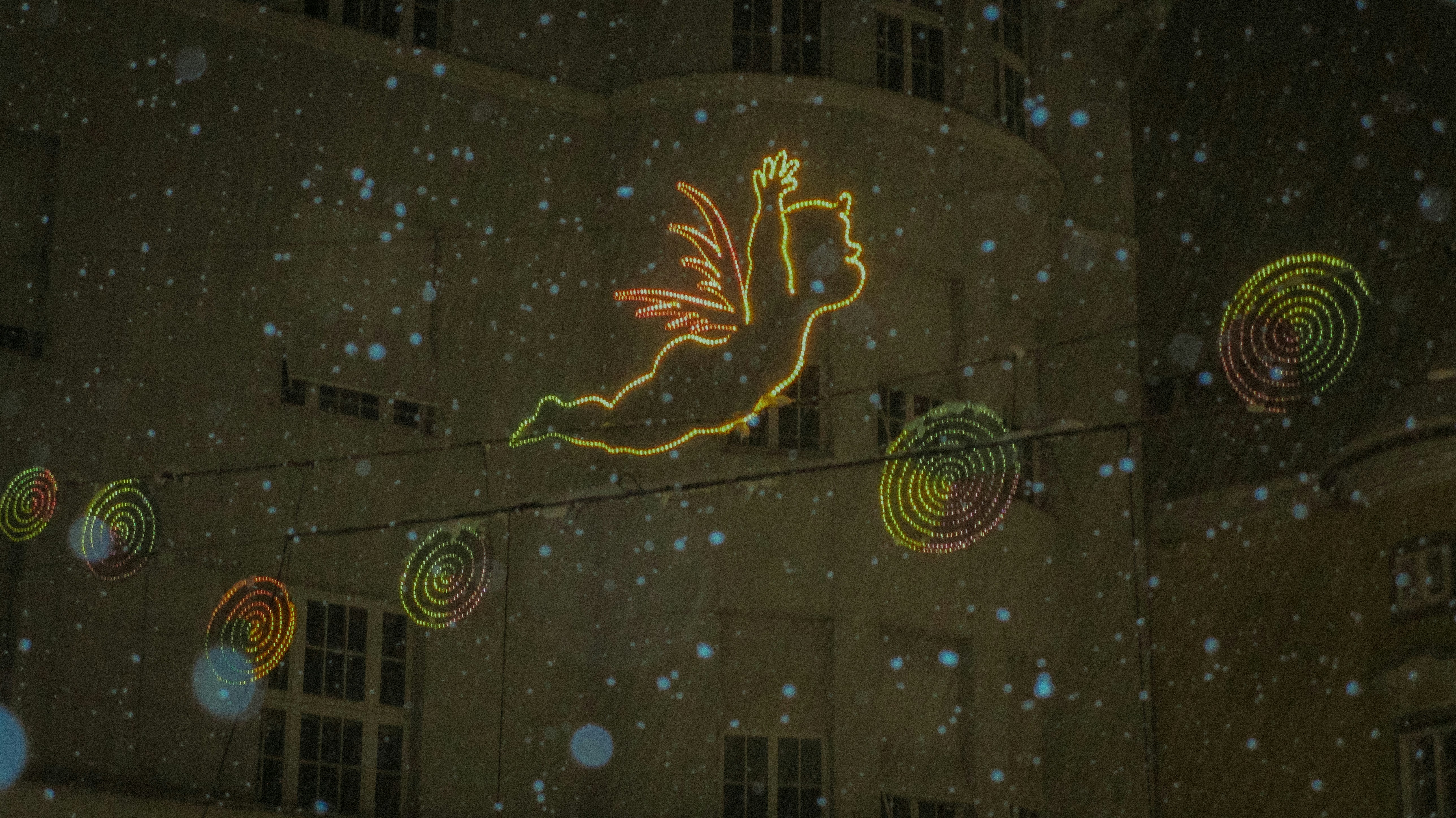Neon angel with wings glows amidst colorful hanging lanterns and falling snow at night.