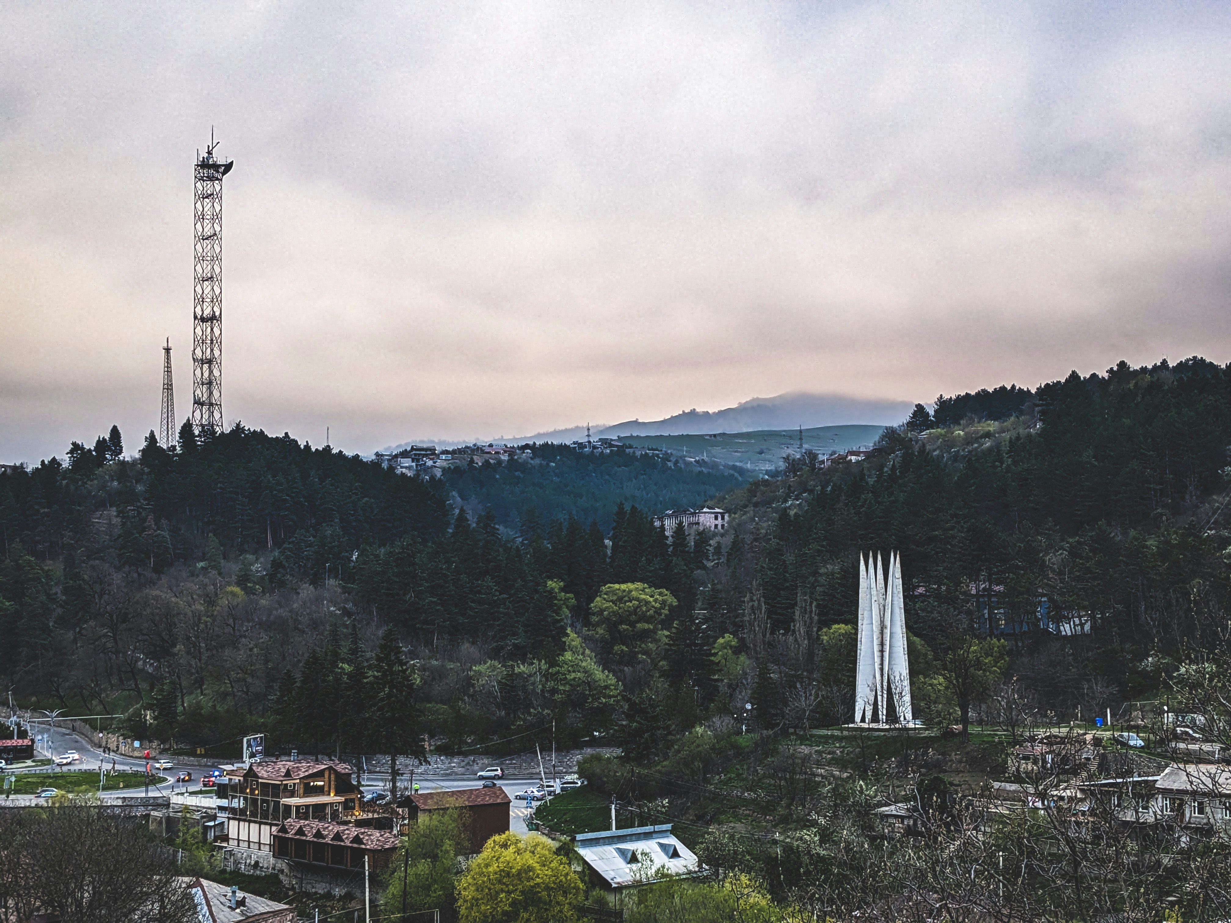 Photograph of a hillside town with a white sculptural sail standing among pines. A tall radio tower anchors the left foreground while low buildings cluster at the base.