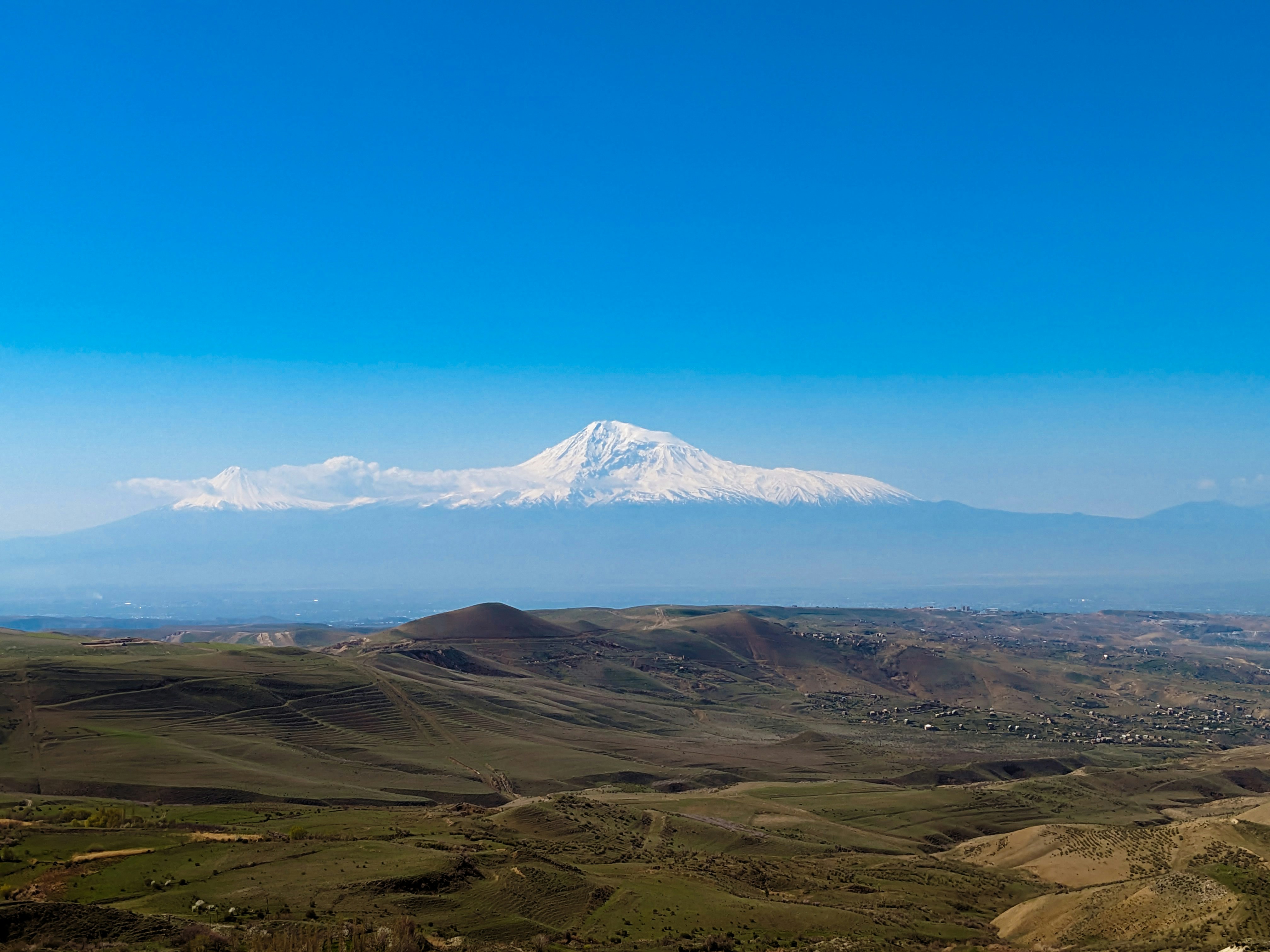 Snow-capped mountain rises above rolling green hills under a crisp blue sky. The wide landscape emphasizes depth and distance in a quiet, natural setting.
