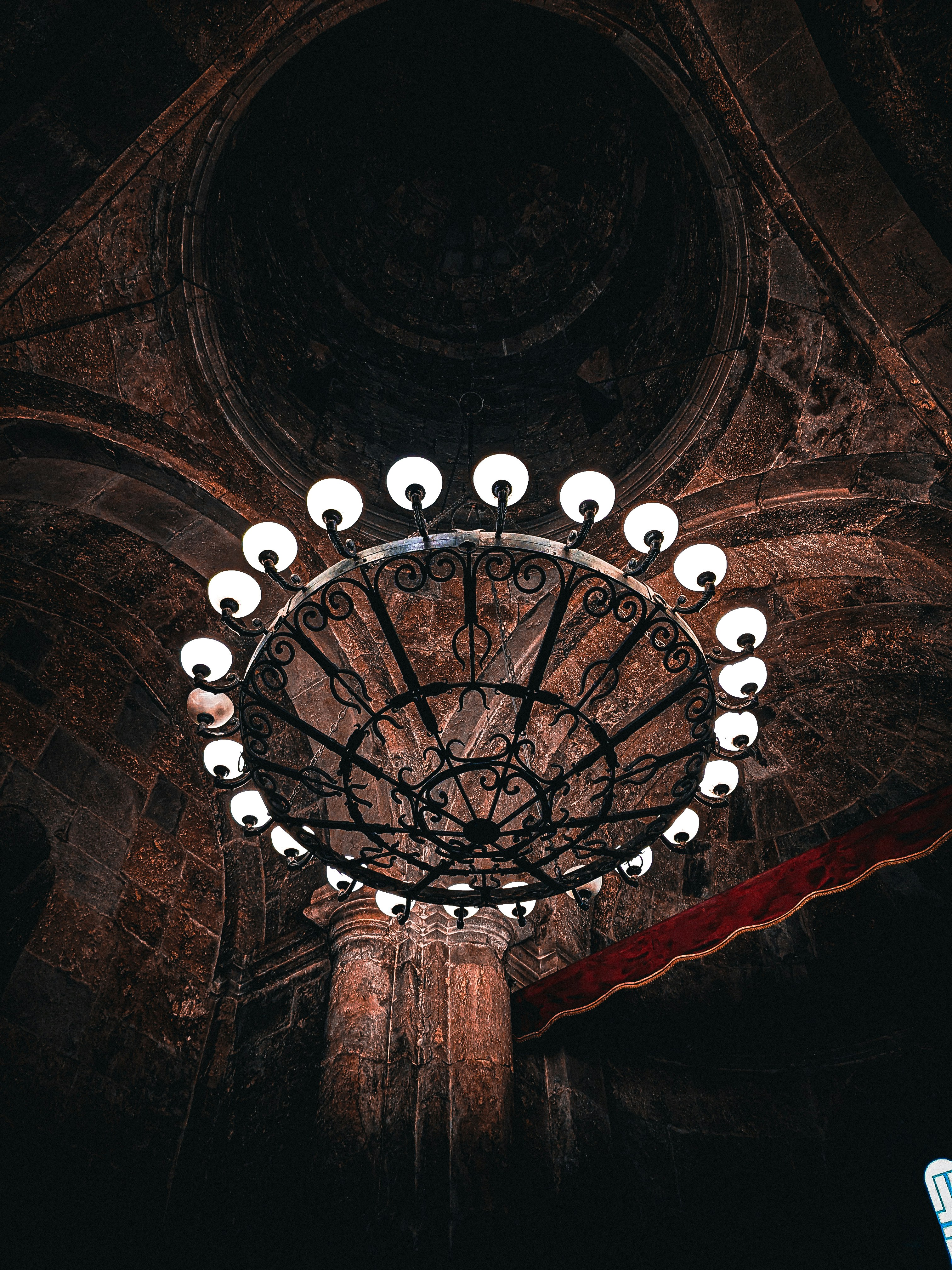 Circular iron chandelier hangs from a dark vaulted ceiling, its ring of bulbs casting warm light over carved stone.
