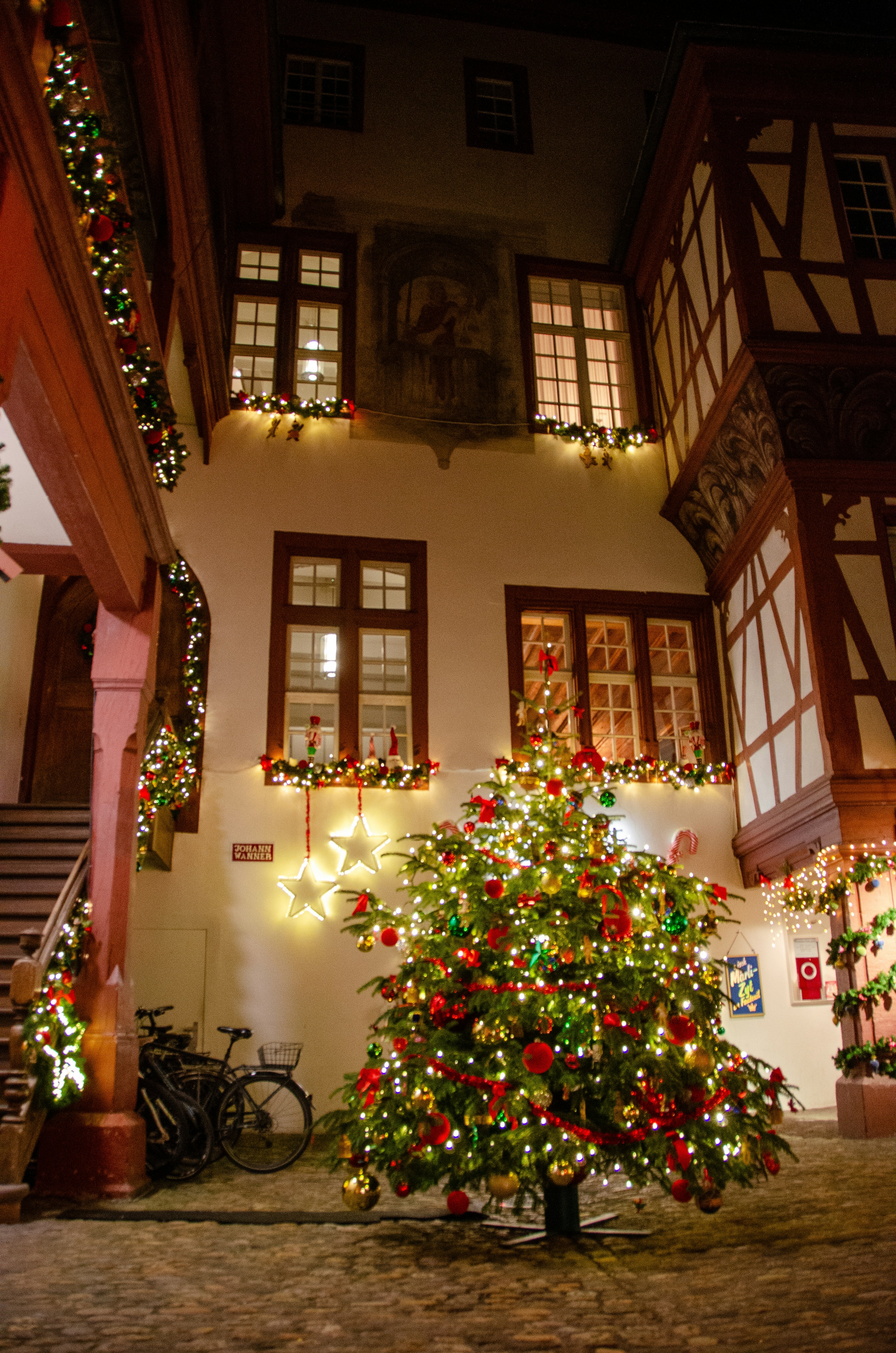 A beautifully decorated Christmas tree adorned with lights and ornaments stands in a charming courtyard, surrounded by festive decorations and historic architecture.