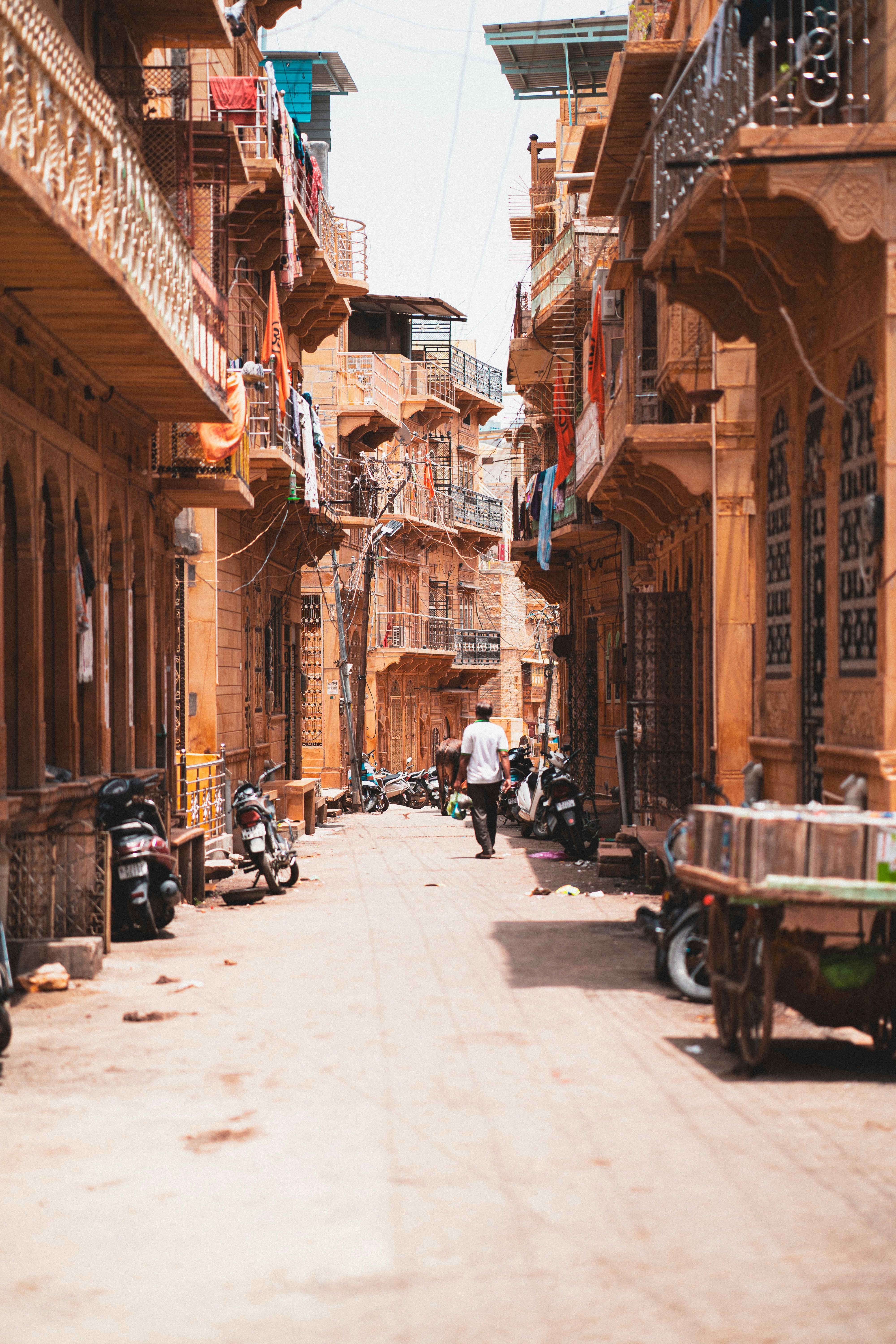 Narrow alleyway in Jaisalmer, showcasing sandstone architecture and a solitary figure walking amidst parked scooters. Bright sunlight illuminates the vibrant colors of the buildings.