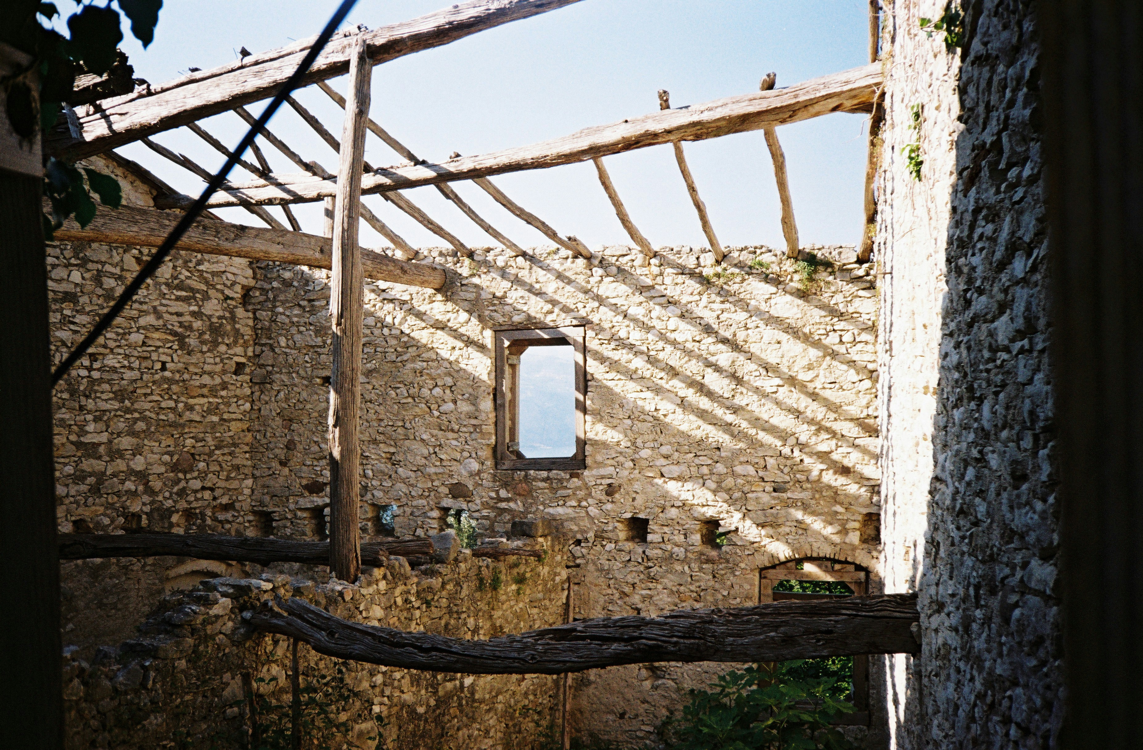 Sunlit interior of a stone ruin with weathered timber beams casting diagonal shadows across the walls. An open window frames a bright sky.