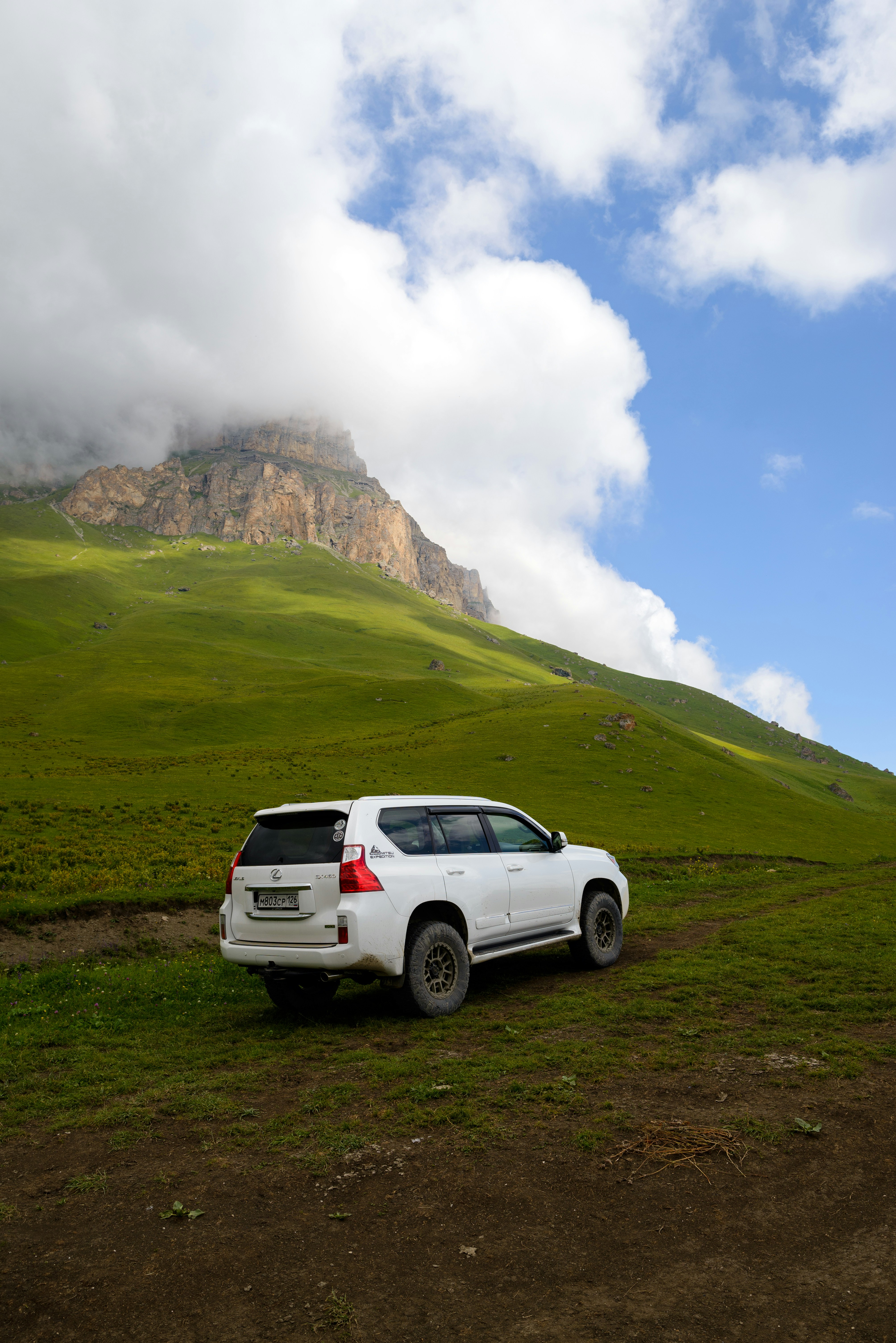 White SUV Climbing a Verdant Ridge Beneath Cloud-Washed Peaks