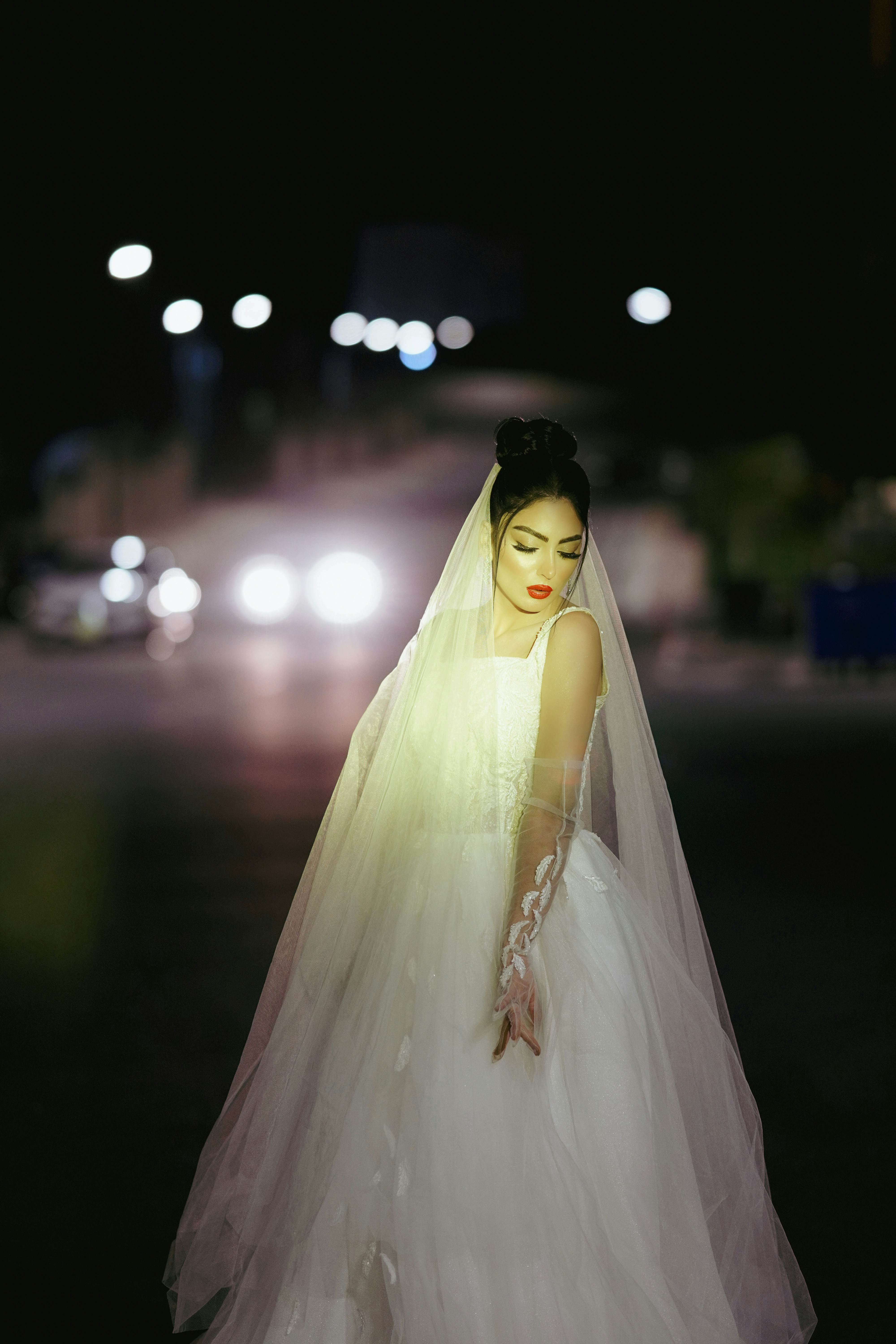 Night portrait of a bride in a white wedding gown with a veil, standing on a dimly lit street. Soft city lights form bokeh behind her as she gazes downward.