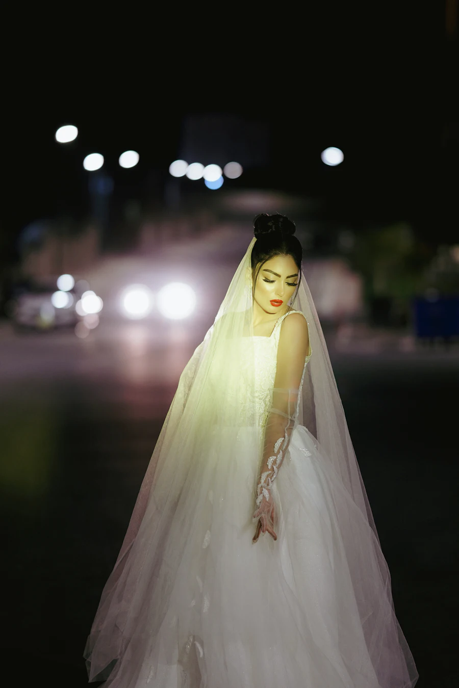 A bride in a white gown and veil stands on a dimly lit street at night, with soft car lights and bokeh glowing behind her. Her face is lowered in a quiet, elegant pose.