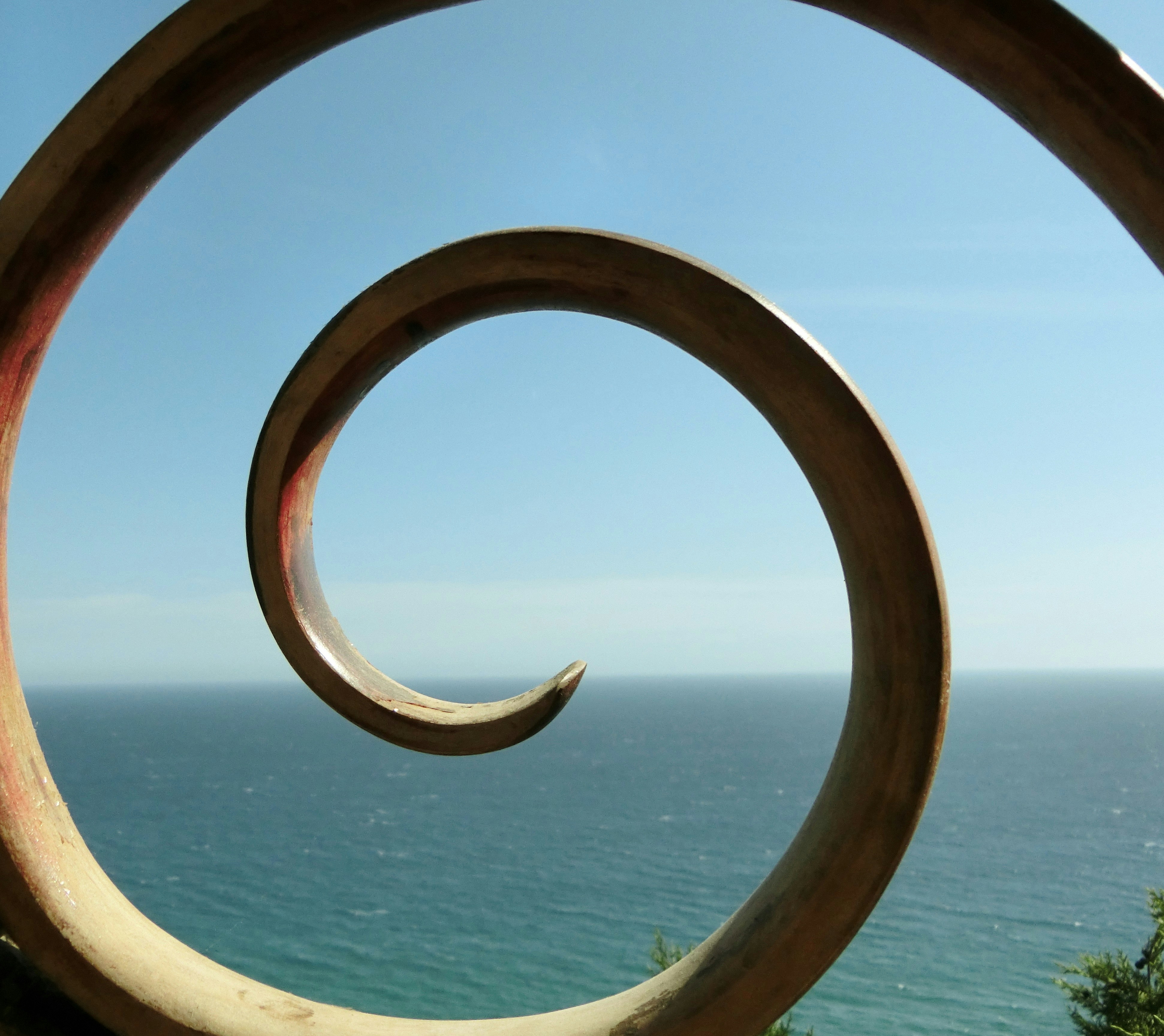 Metal spiral sculpture framing a vast ocean horizon under a clear blue sky.