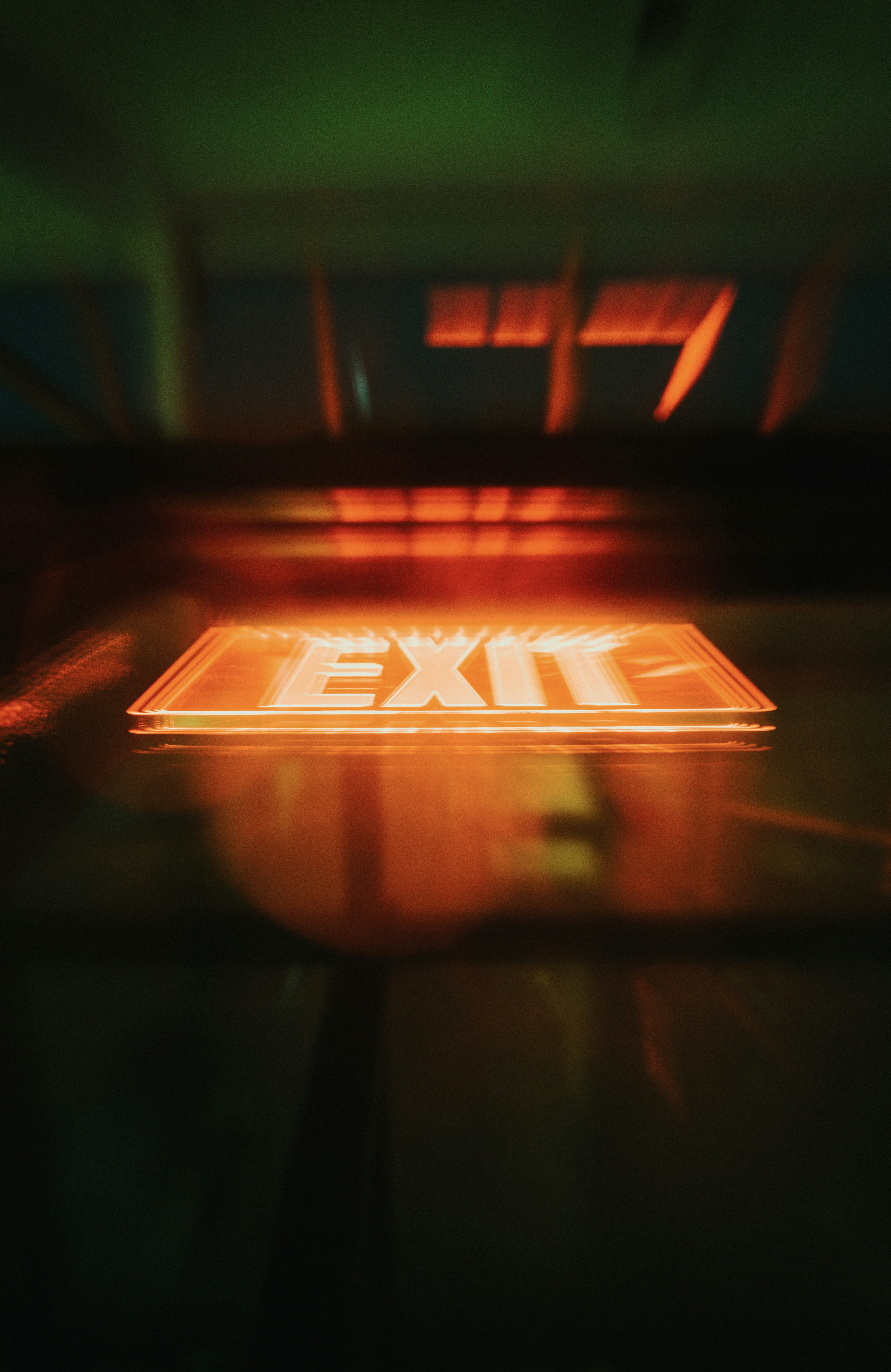A vibrant red exit sign is viewed through a window, creating a surreal and abstract visual effect. The image is dominated by deep reds and contrasting dark shadows, with soft, diffused lighting that adds a mysterious atmosphere. The composition draws the viewer's eye to the bold, glowing text, making the sign appear almost ethereal against the darker surroundings.