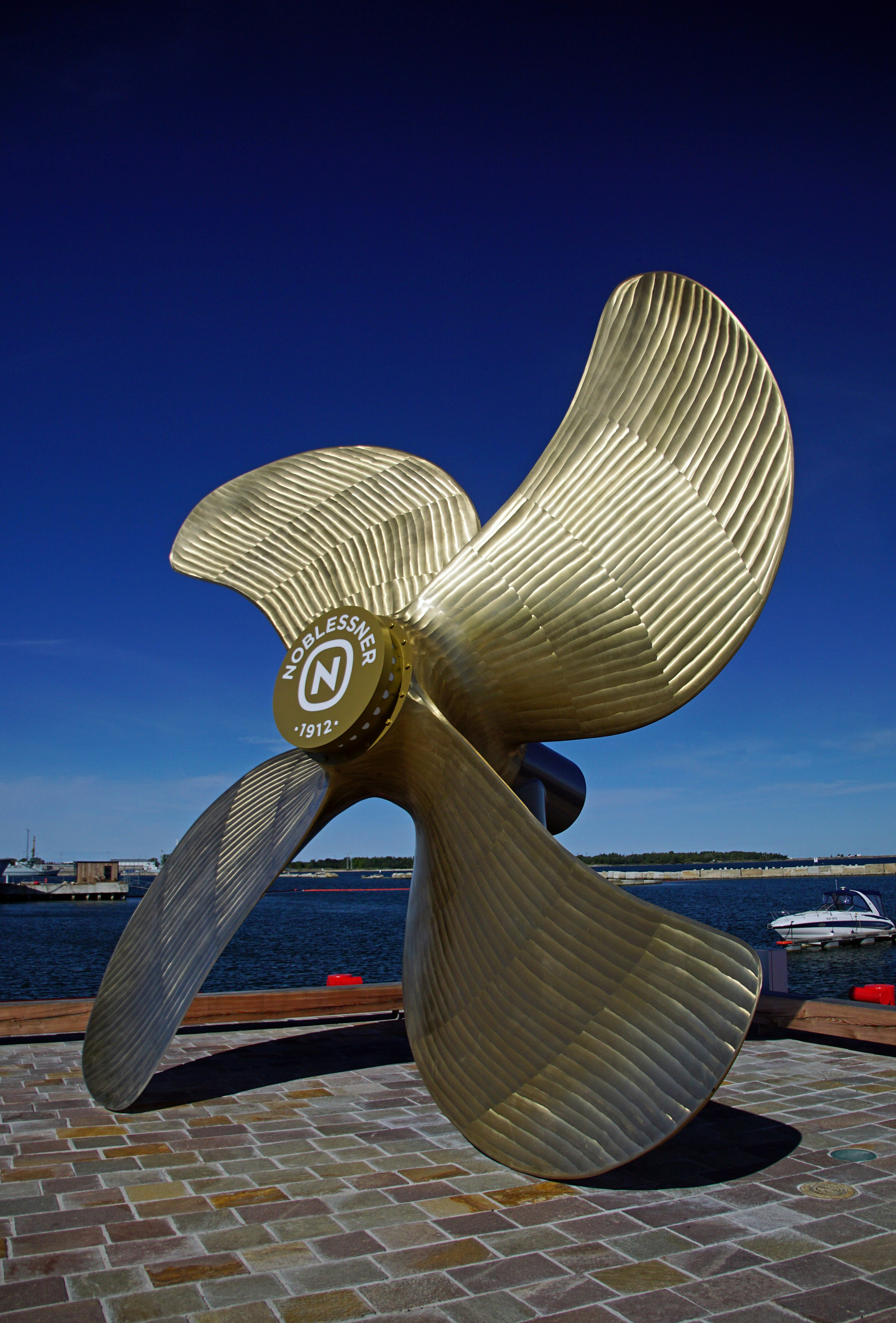 A large, golden boat propeller on display in the harbor of Tallinn in Estonia on a sunny summer day. | a large metal propeller sitting on top of a brick walkway
