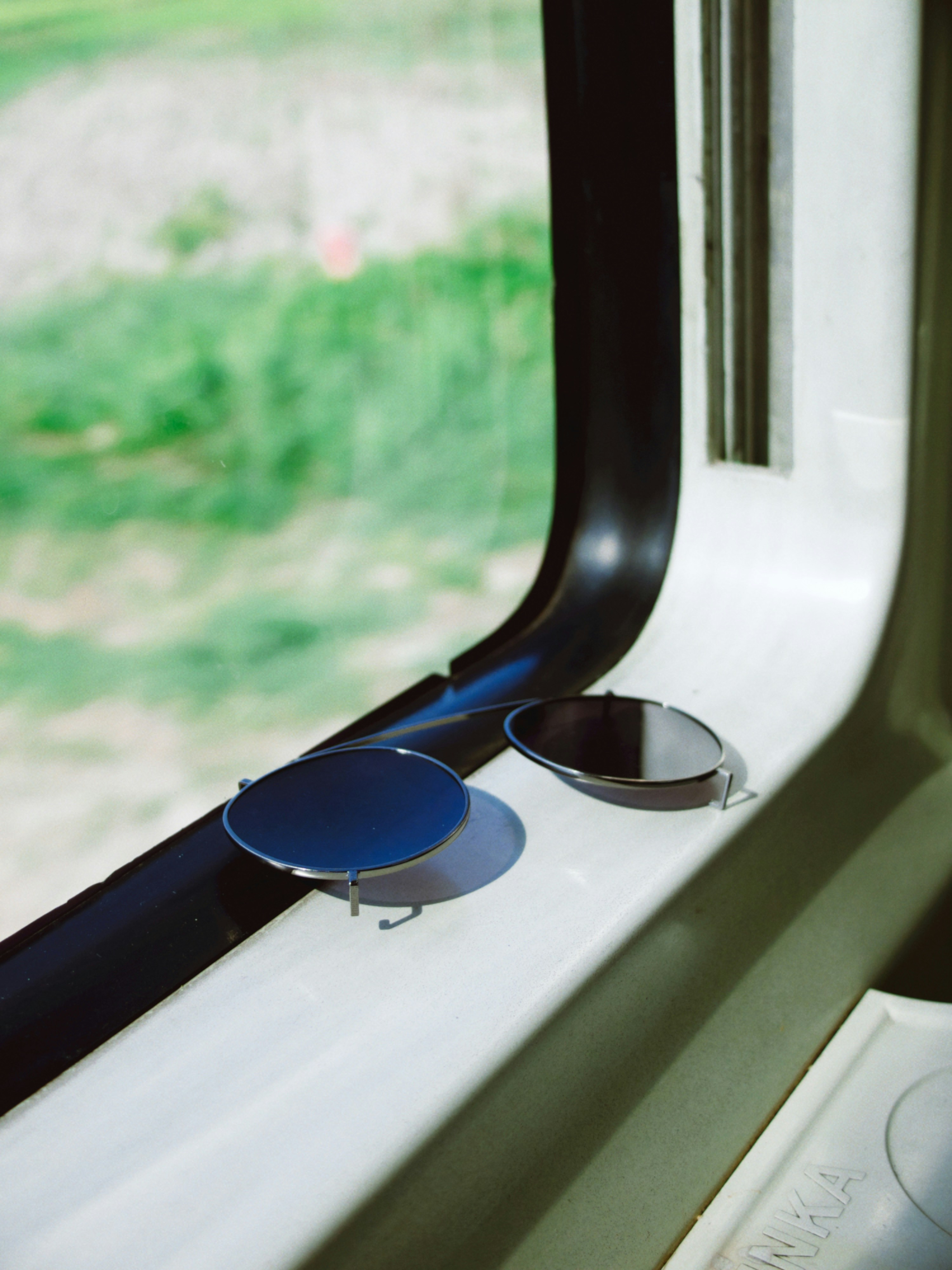 Close-up of round blue sunglasses resting on a sunlit windowsill with a softly blurred green landscape outside.
