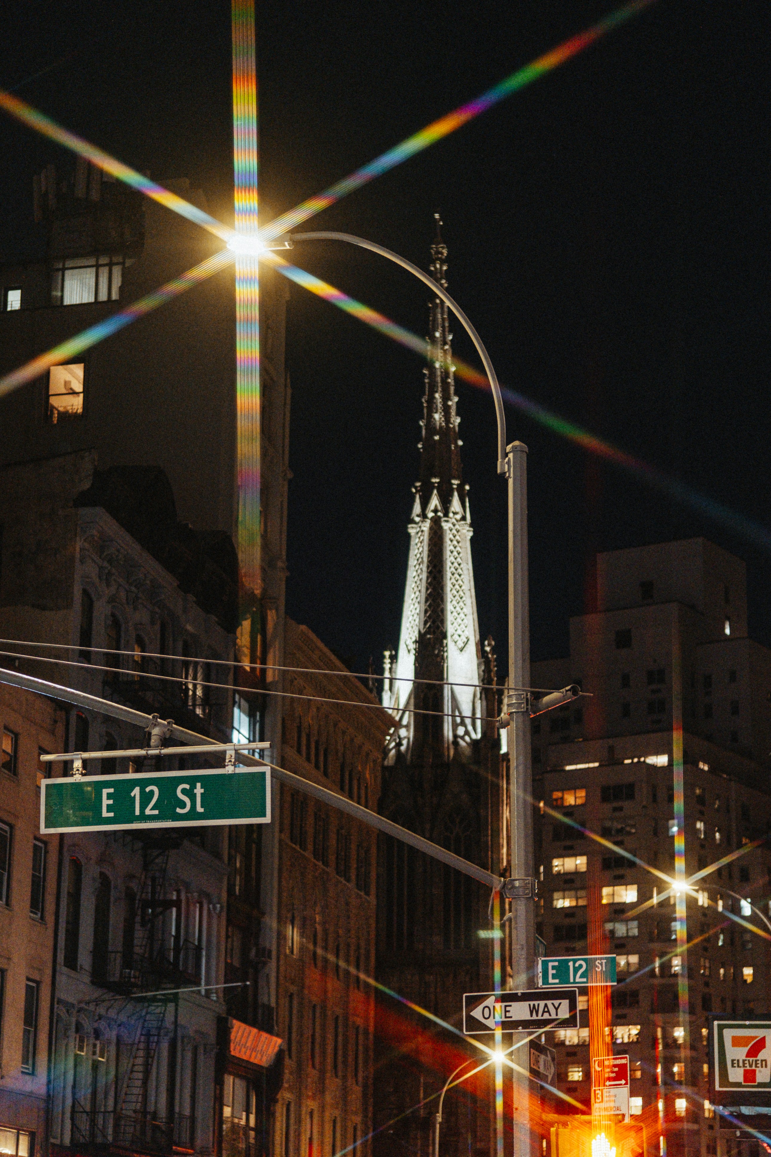 Nighttime urban street photograph with rainbow diffraction from streetlights across signs and a towering Gothic spire in the background.