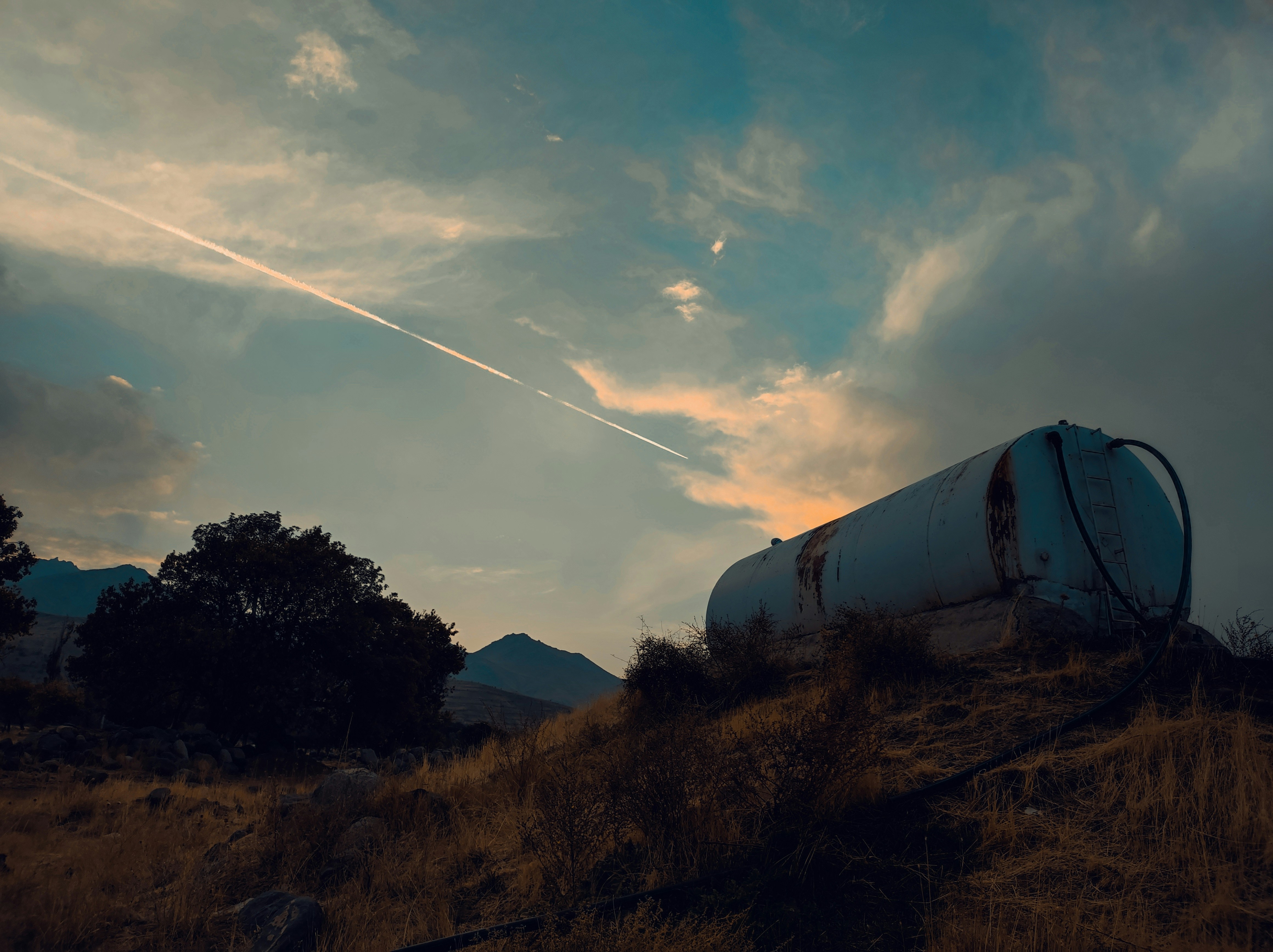 A weathered tank rests on a hillside, surrounded by sparse vegetation and distant mountains under a moody sky. Contrails streak across the twilight atmosphere.
