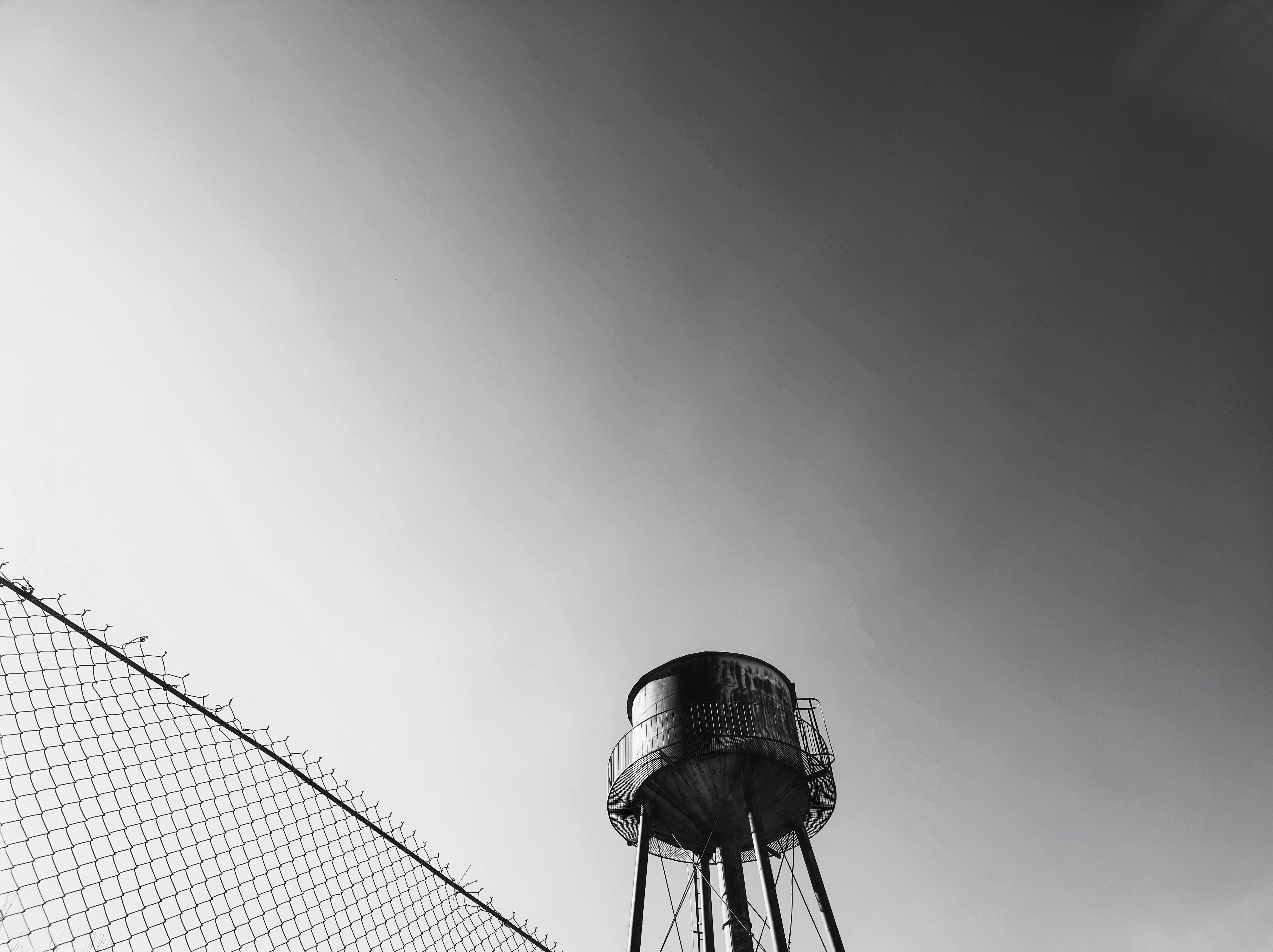 Monochrome photograph of a water tower on slender legs against a diagonal chain-link fence. The stark gradient sky emphasizes geometry and texture.