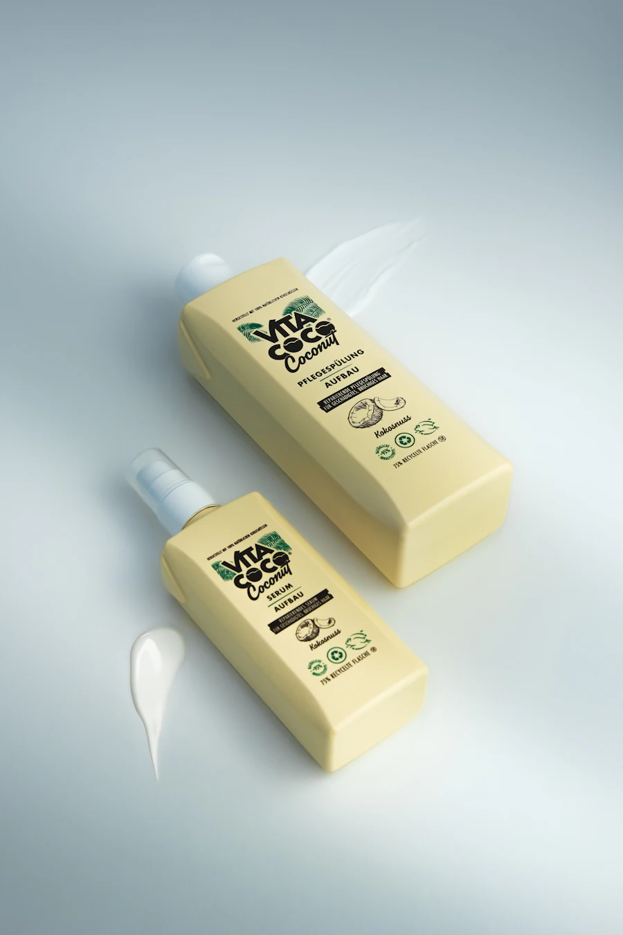 Two pale yellow Vita Coco skincare bottles with white caps lie diagonally on a soft light-blue surface, with small white cream swirls nearby. Minimal product styling with plenty of negative space.