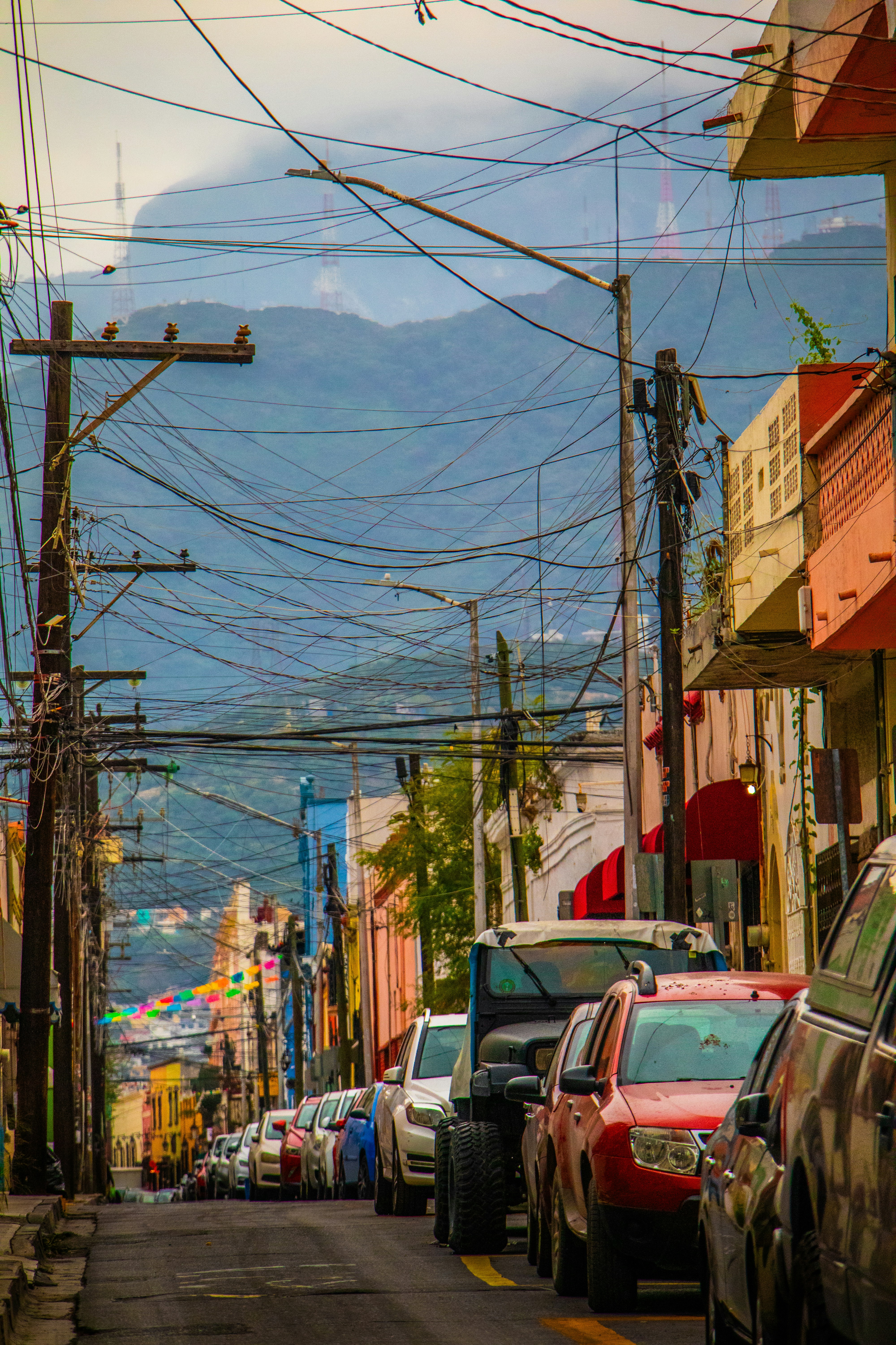 Colorful cars line a narrow street beneath a tangle of power lines, with mountains in the distance.