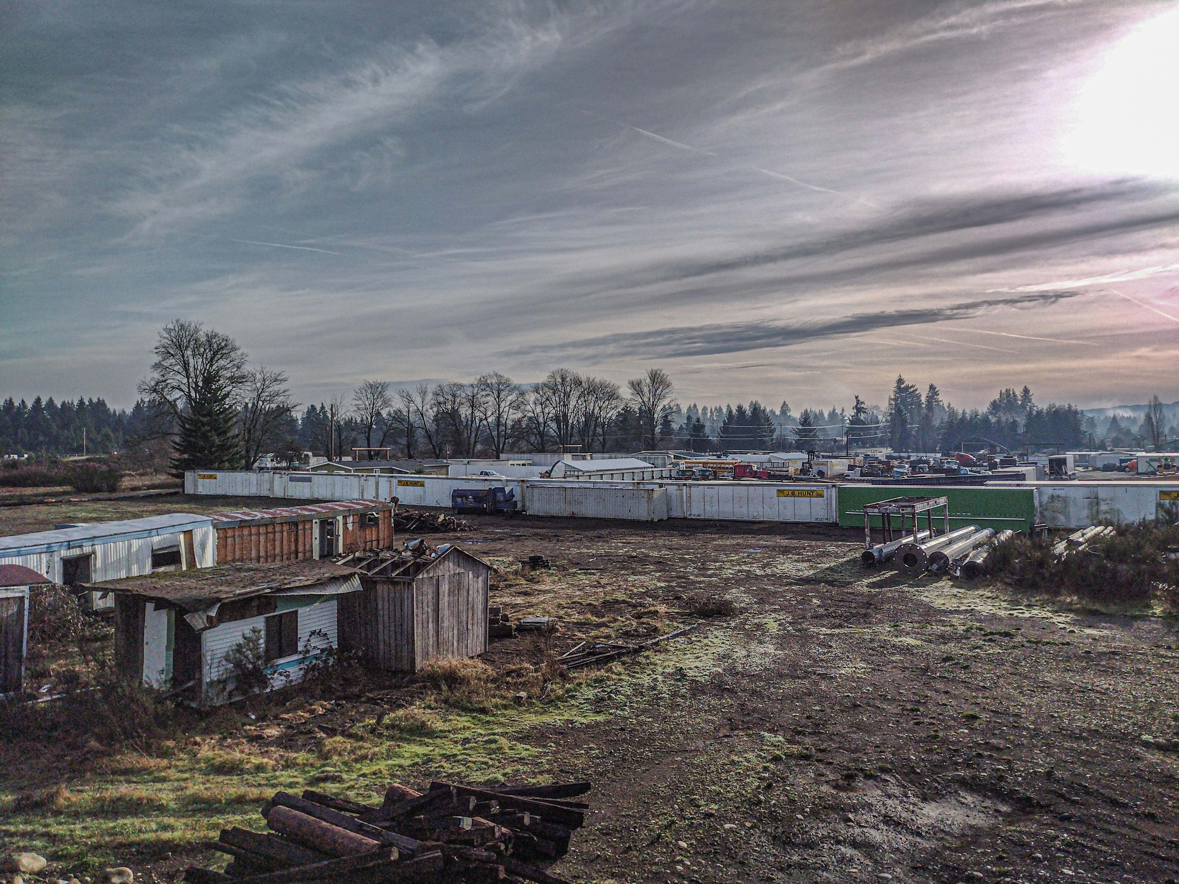 Landscape photograph showing a weathered yard with old sheds in the foreground and a row of storage huts on the horizon. Rugged textures contrast with the open sky.