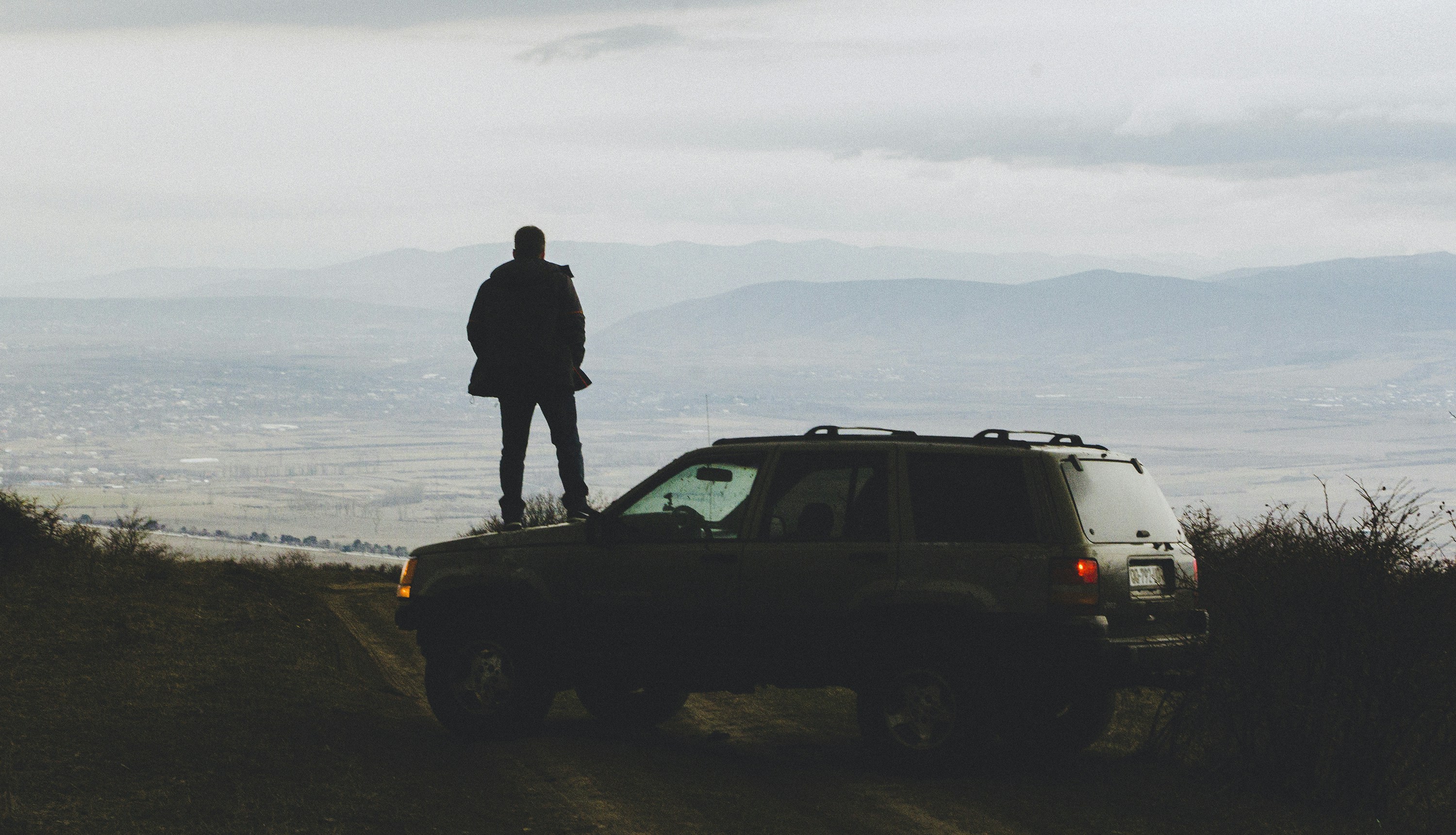 Silhouette of a person standing on a vehicle, overlooking a vast landscape under a cloudy sky. The scene conveys a sense of adventure and contemplation.