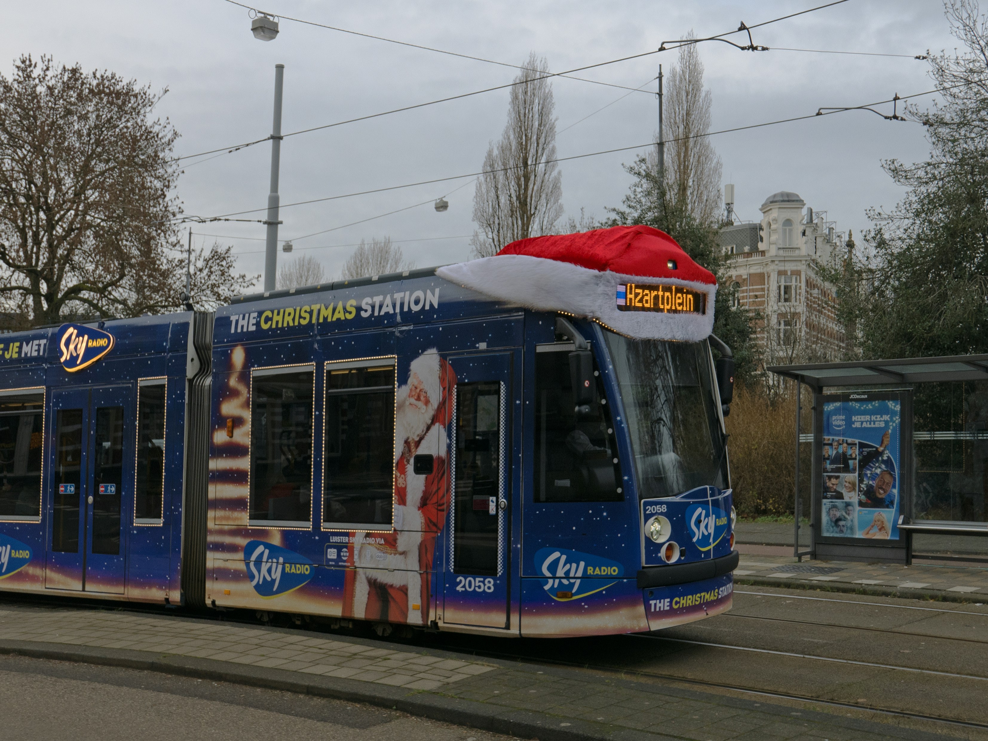 Free photo Amsterdam with Christmas tram with red hood / hat / cap - driving in the street Sarphatistraat, near the tram stop. It is grey weather in early winter. Urban street photography in free images - high resolution and free download for poster, print & wallpaper - street photos of cities in The Netherlands by Fons Heijnsbroek / Gratis foto van de blauwe Kersttram met rode muts voorop. Hij rijdt in Amsterdam over de trambaan op Sarphatistraat. Gratis download foto - straatfotografie van stad in Nederland, fotograaf Fons Heijnsbroek