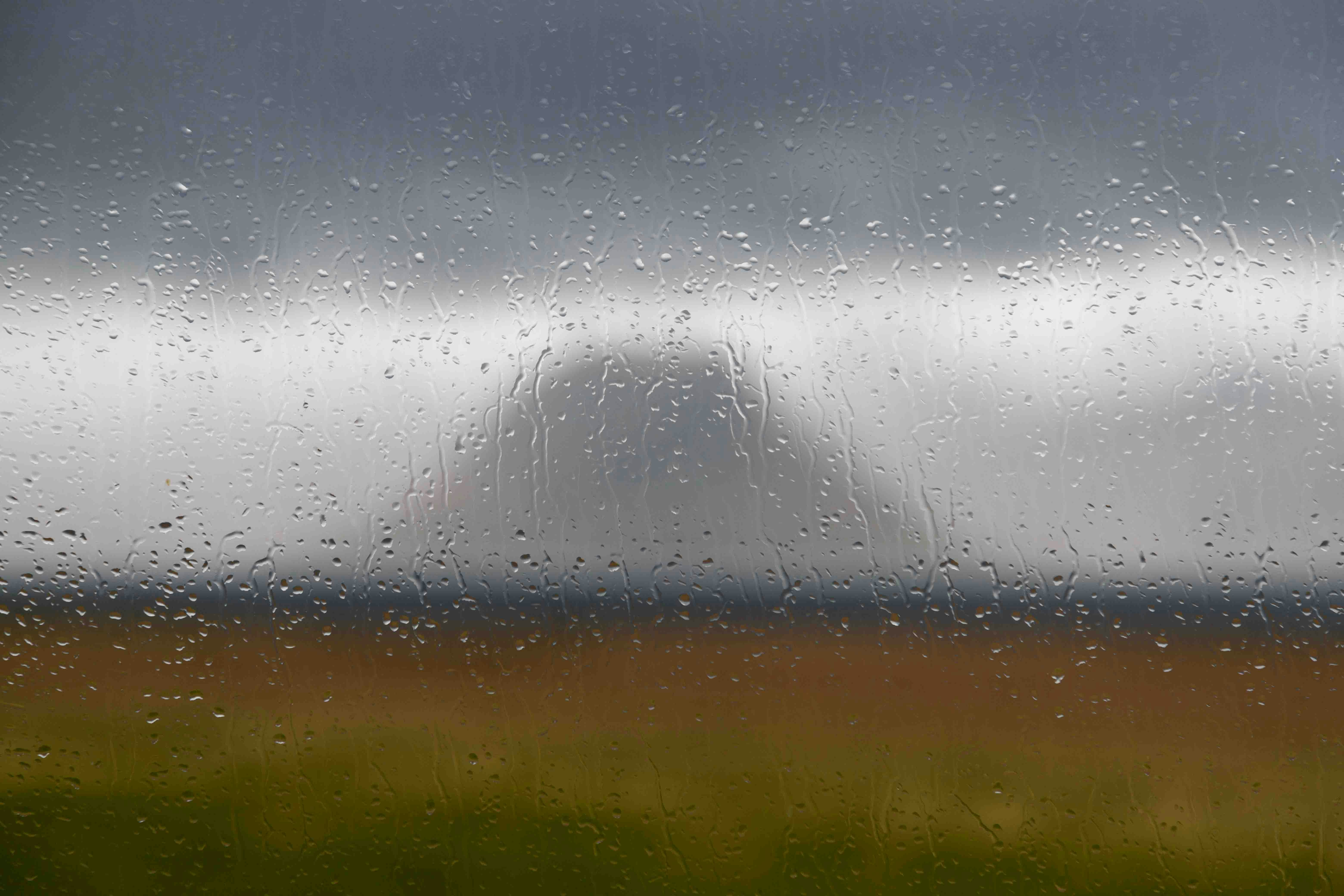View of a rainy landscape through a wet window, with raindrops scattered across the glass, obscuring the details of a field and a mountain in the background.