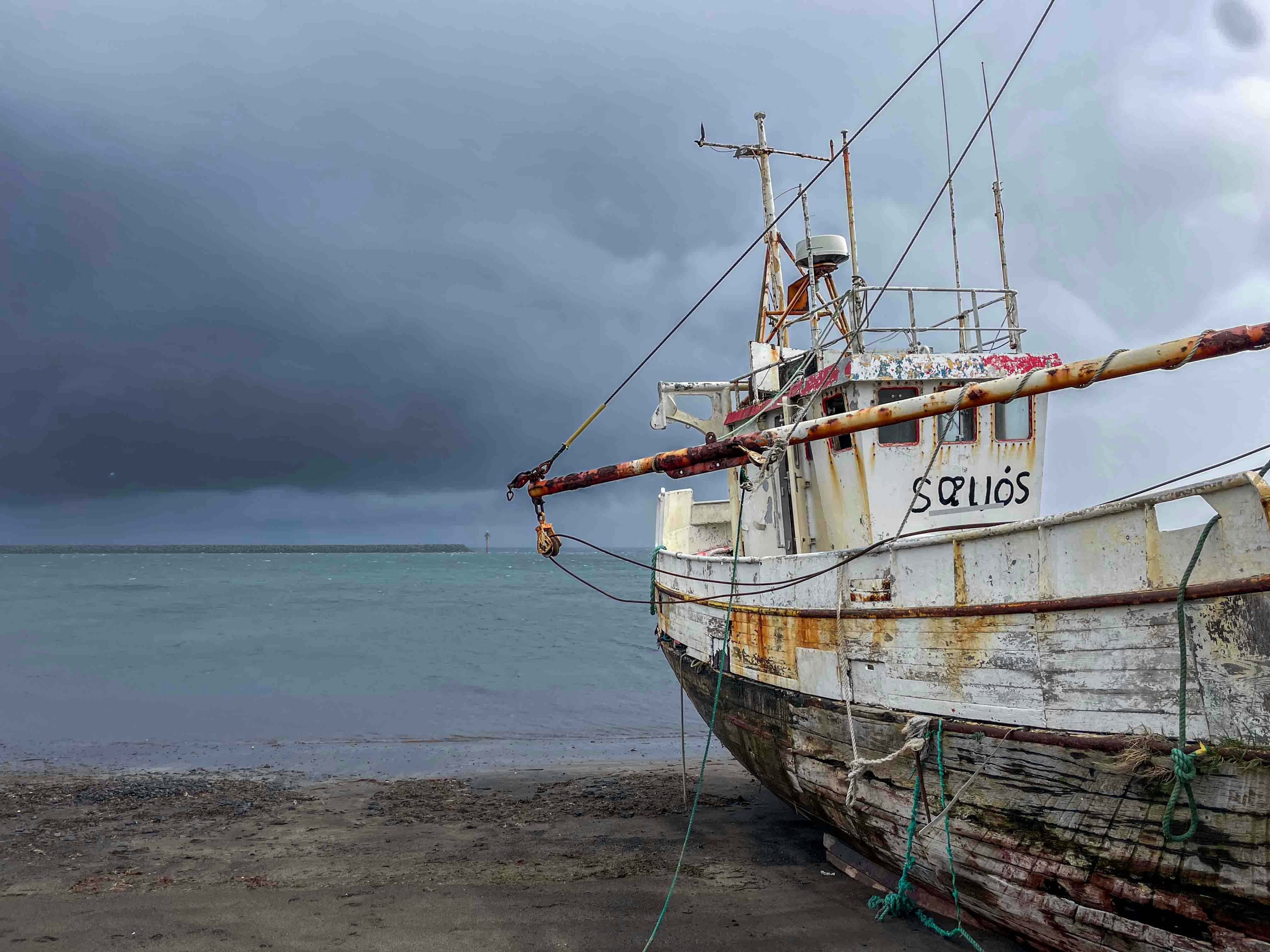 A weathered fishing boat, named Sæliós, beached on a dark sandy shore under a gloomy sky, conveying a sense of abandonment and time's passage.